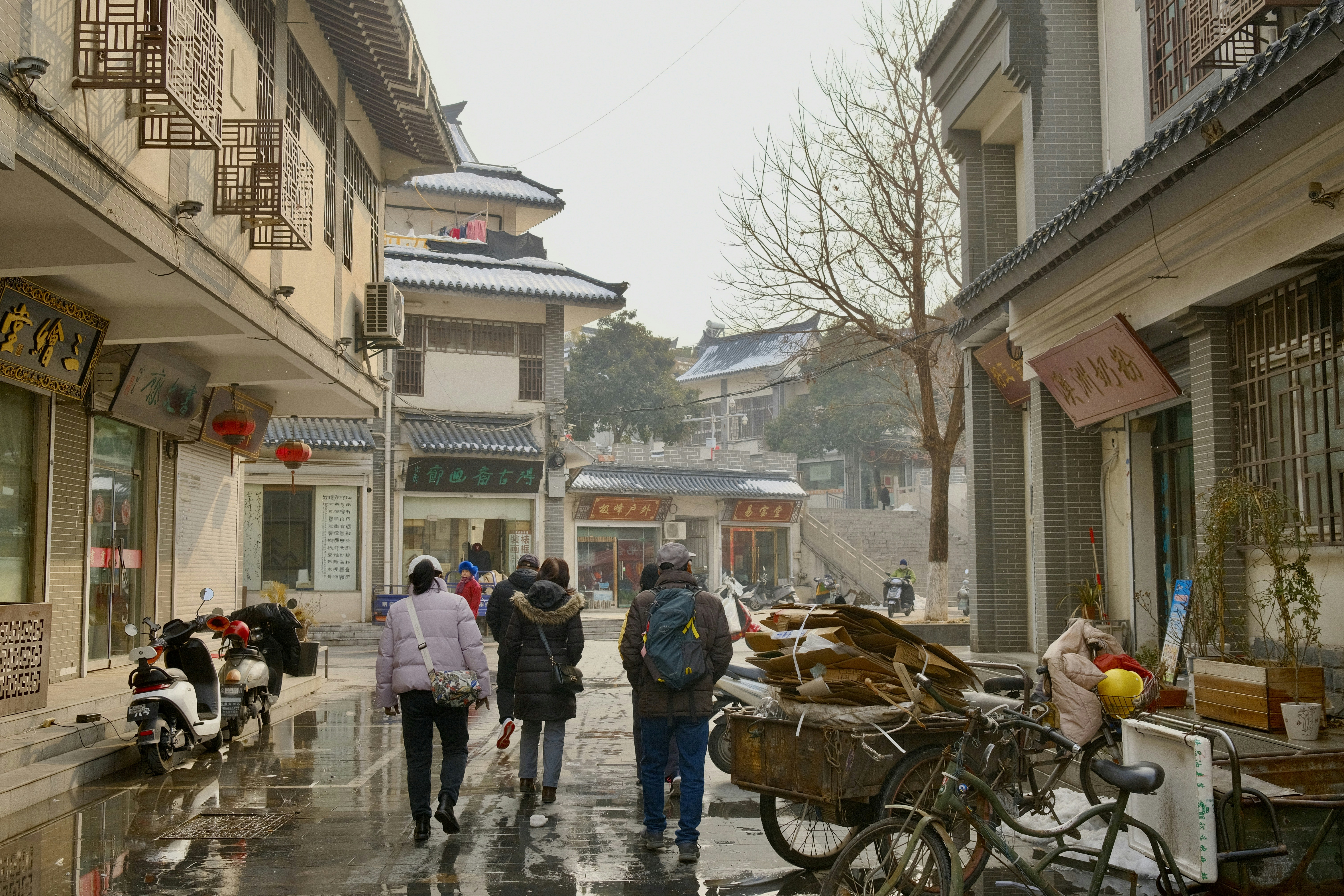 Four people walk down a wet, narrow street lined with traditional buildings and bicycles on a cloudy day.