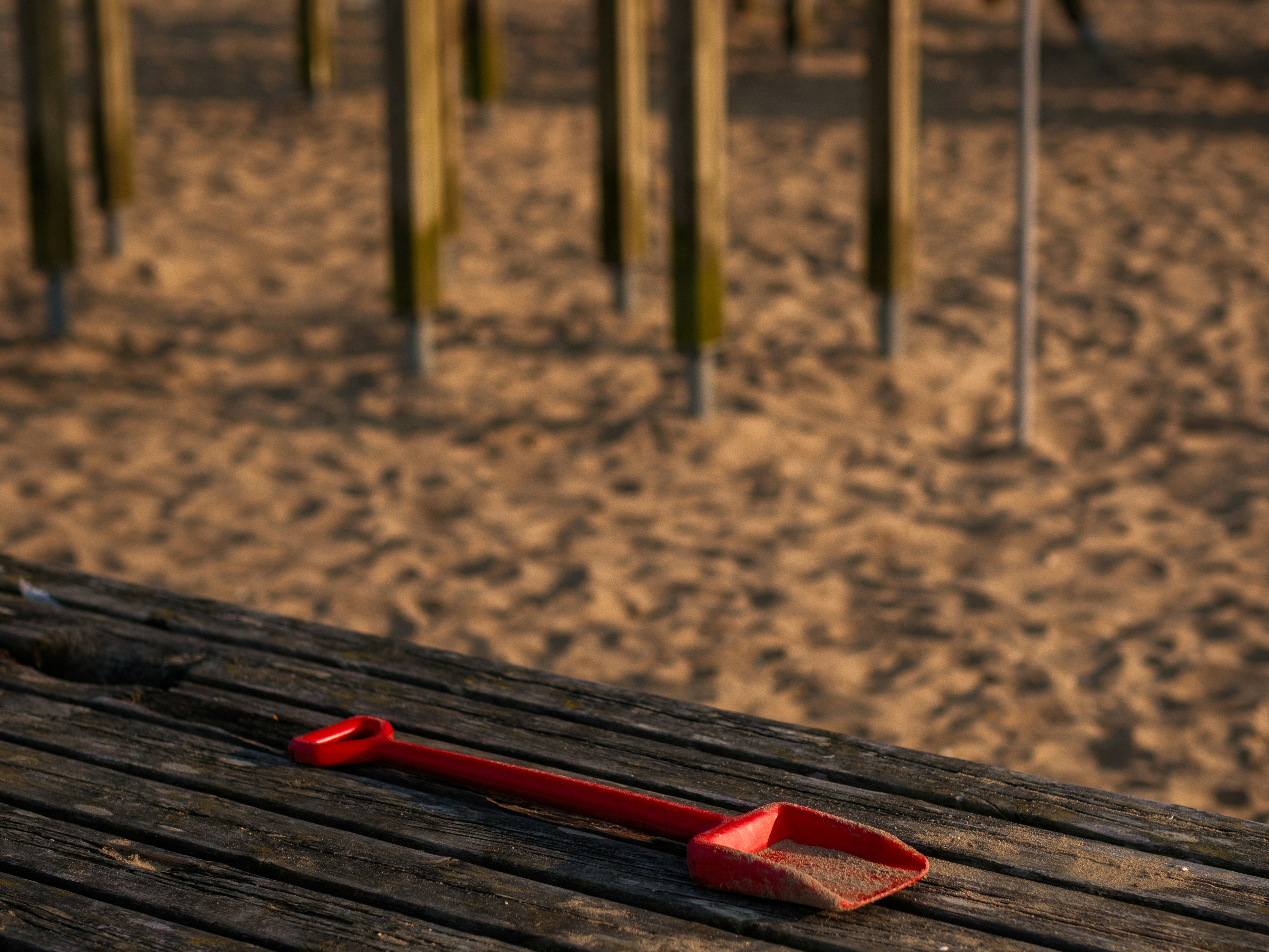 Red shovel resting on a wooden table with a sandy playground backdrop.