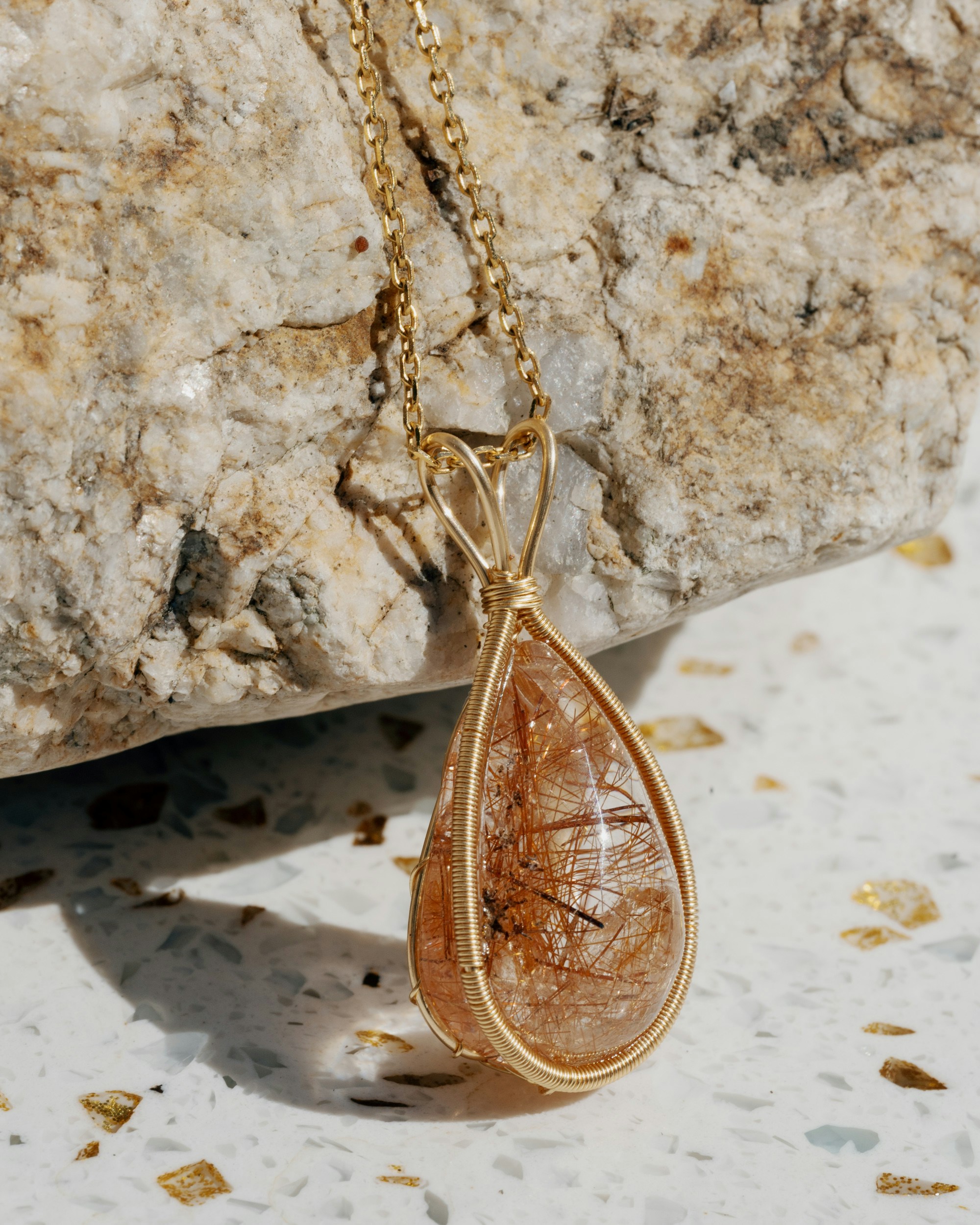 Gilded Teardrop: Rutilated Quartz Pendant Nestled on Rough Stone