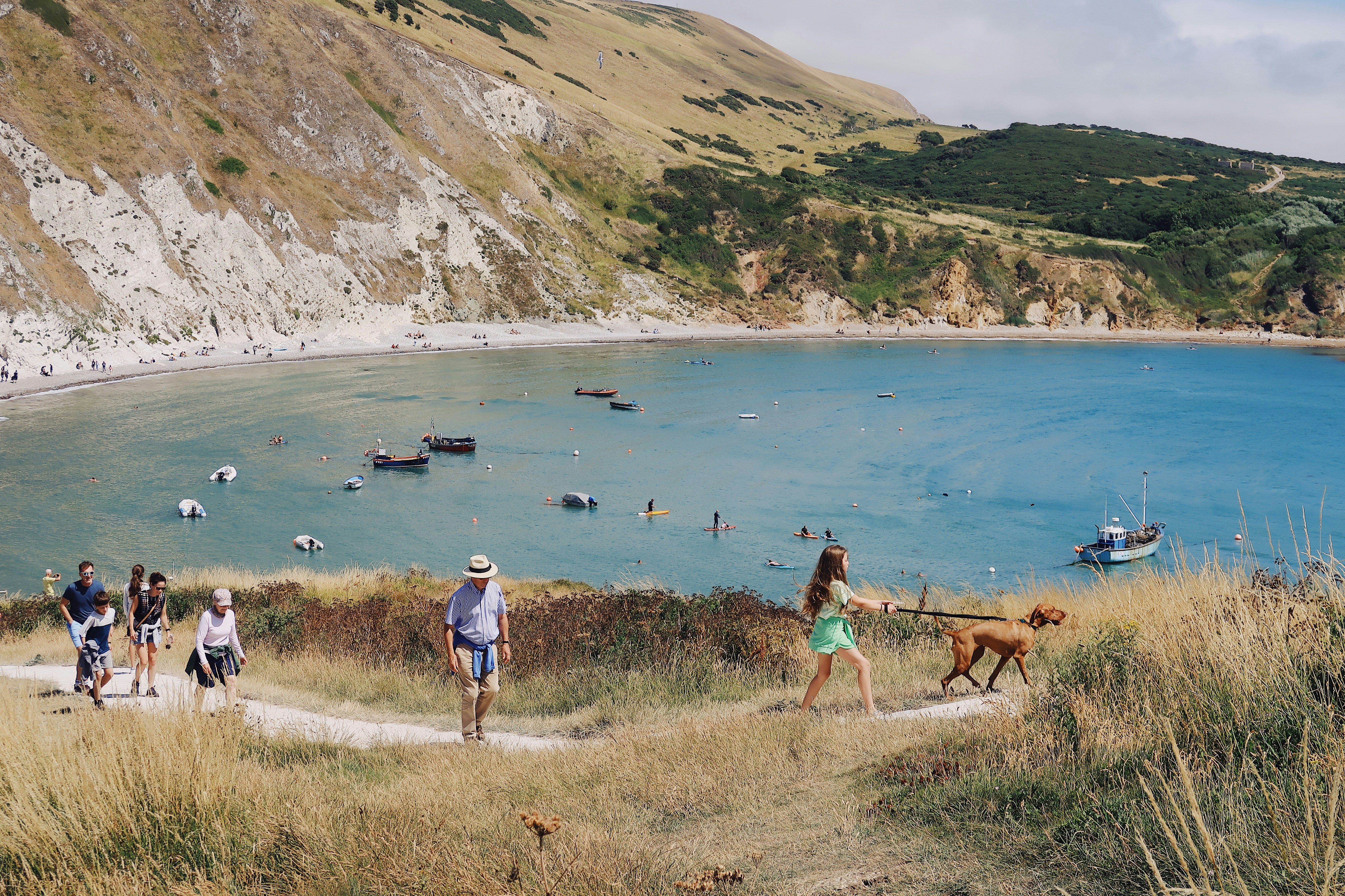 Group of people walking along a coastal path with a dog, overlooking a tranquil bay dotted with boats. The scene captures the essence of a leisurely day outdoors.