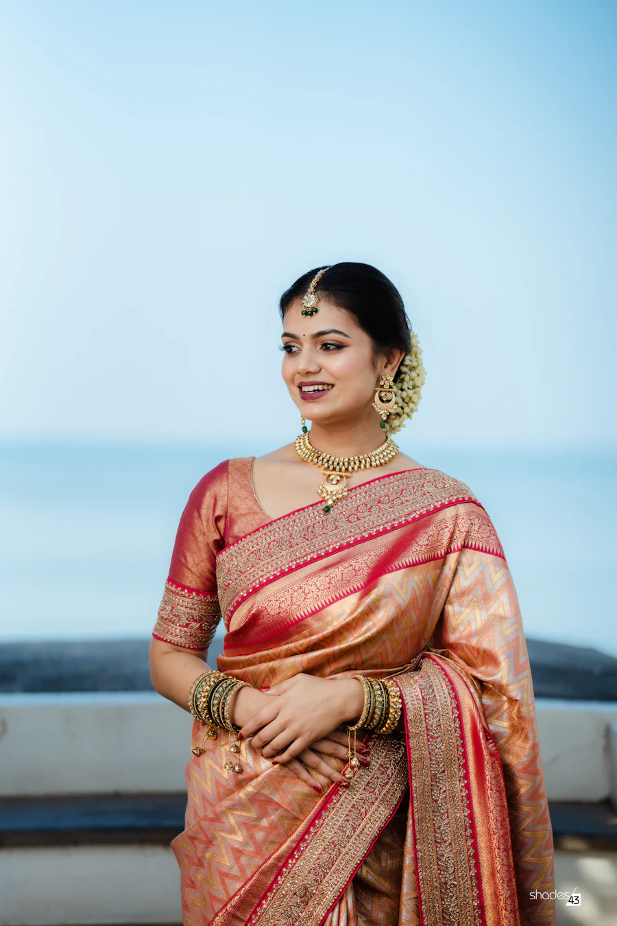 A smiling woman in a pink and gold sari with traditional jewelry stands against a soft blue sea and sky backdrop. Her bangles, necklace, and floral hair adornments add ornate detail.