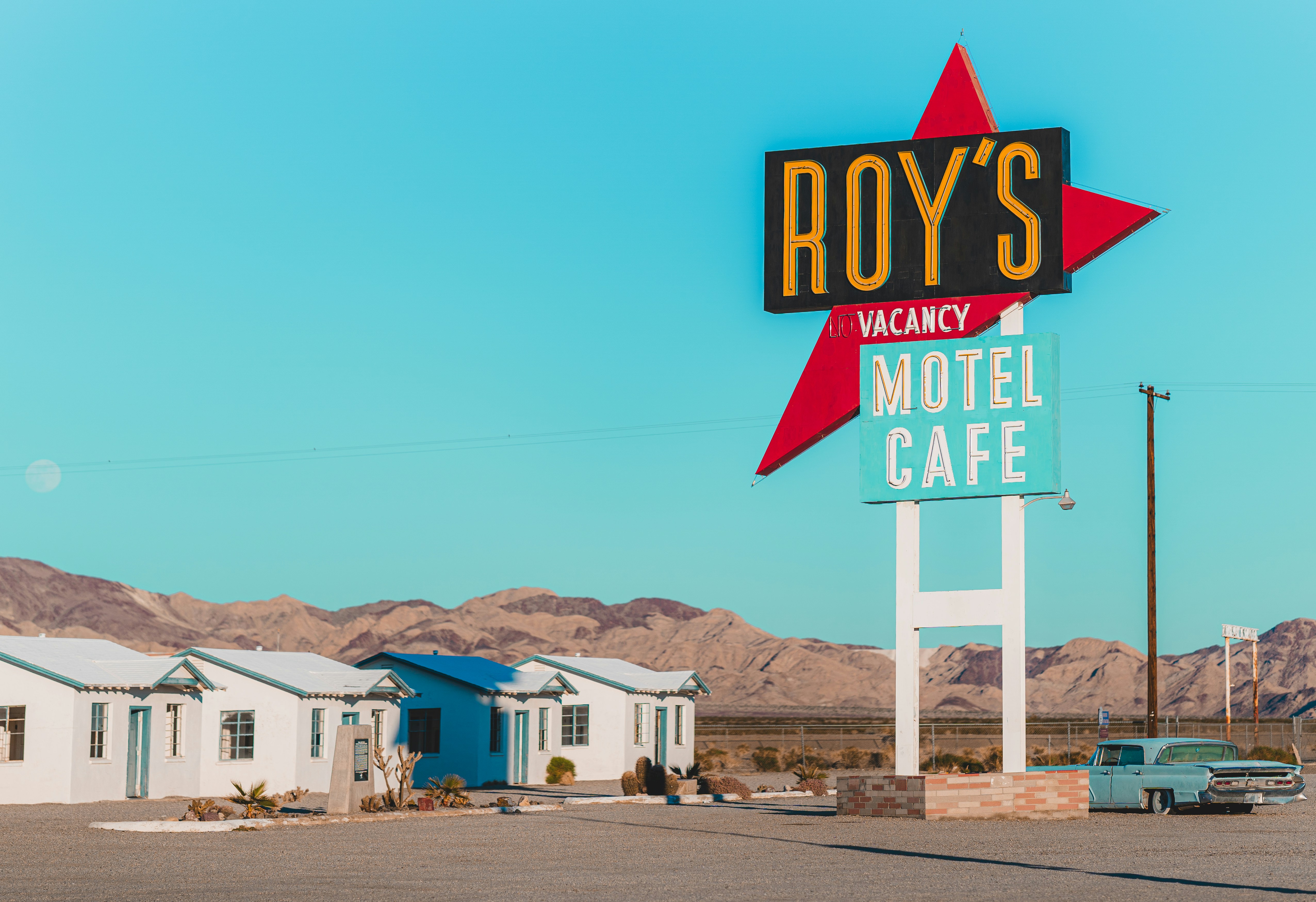 Vibrant vintage motel sign stands against a clear blue sky, with desert mountains in the background and classic cars parked nearby.