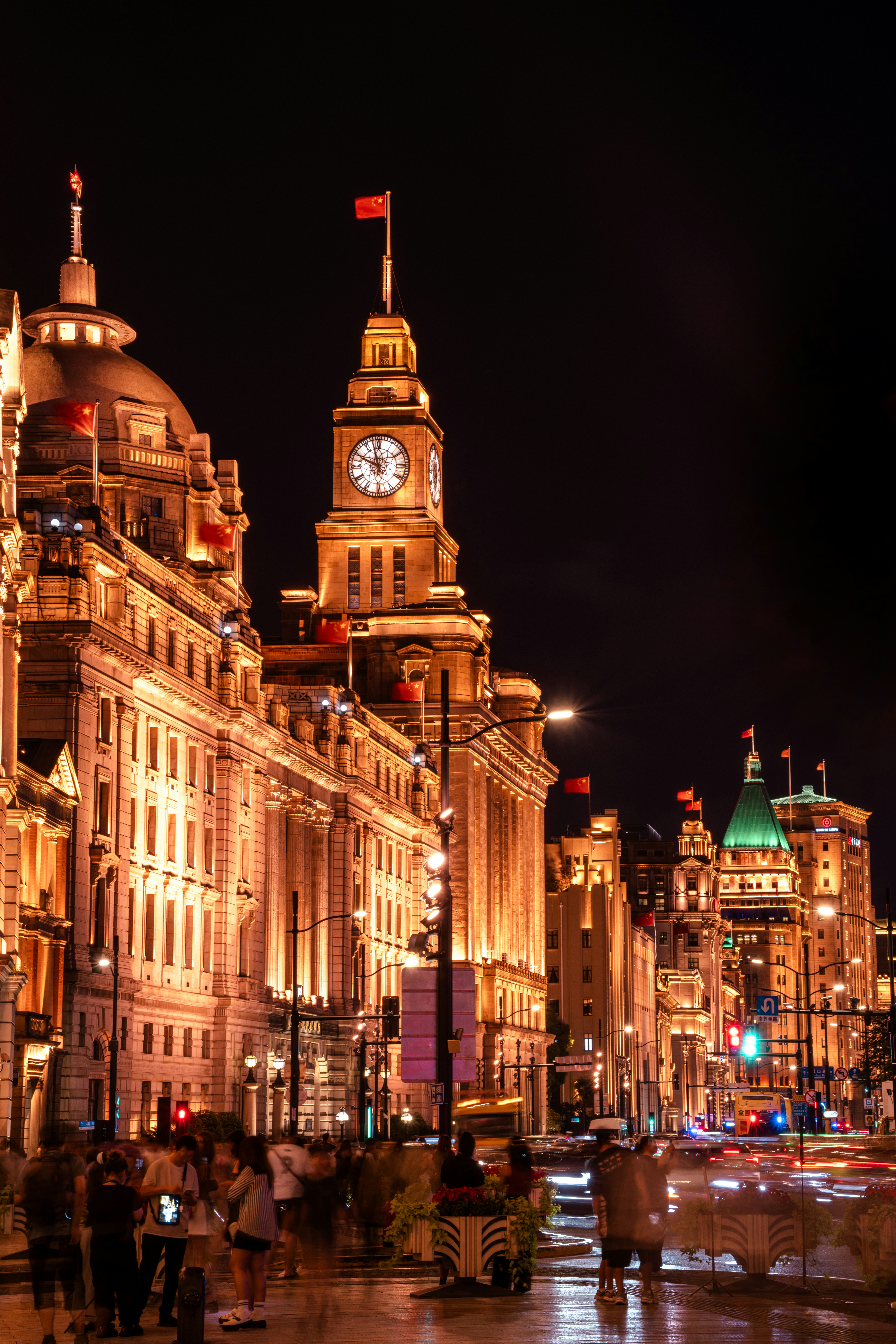 Photograph of a grand Bund street at night, with warm, illuminated historic façades and a prominent clock tower, flags atop roofs, and blurred pedestrians and moving traffic.