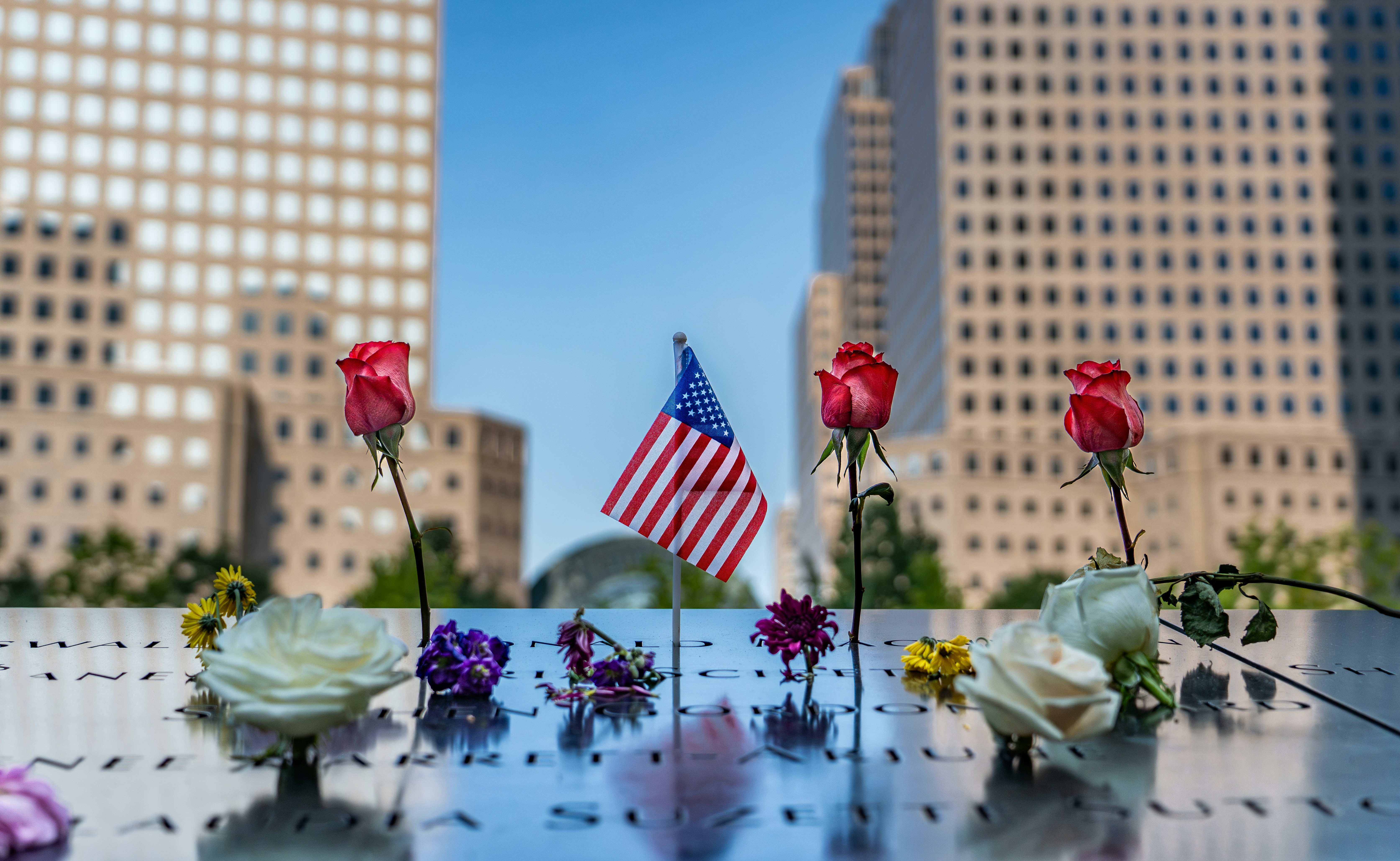 Small American flag and flowers placed at a memorial site, surrounded by towering buildings. The scene reflects remembrance and resilience.