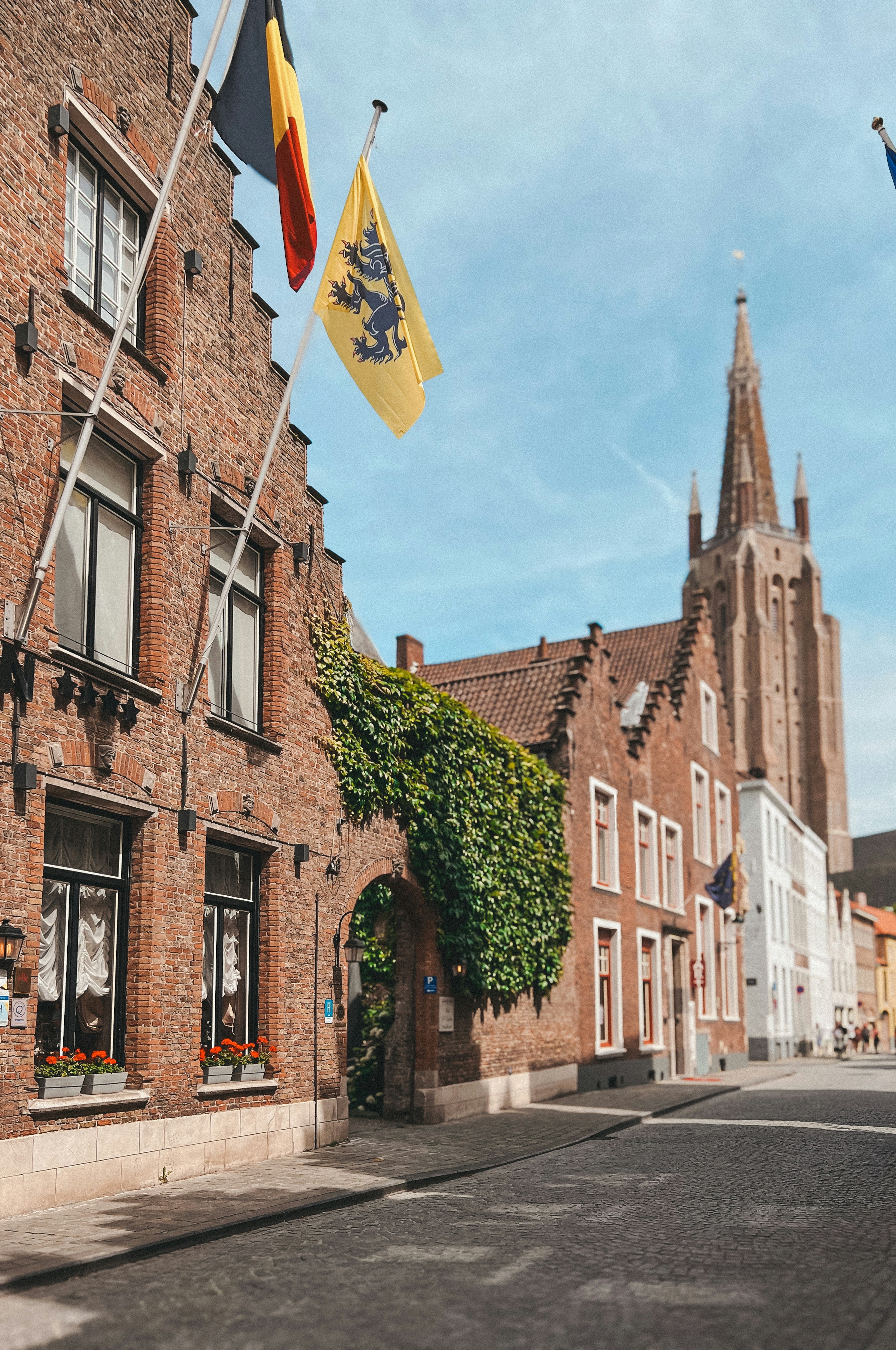 Charming brick buildings adorned with flags and greenery line a cobblestone street, leading to a historic tower in the background.
