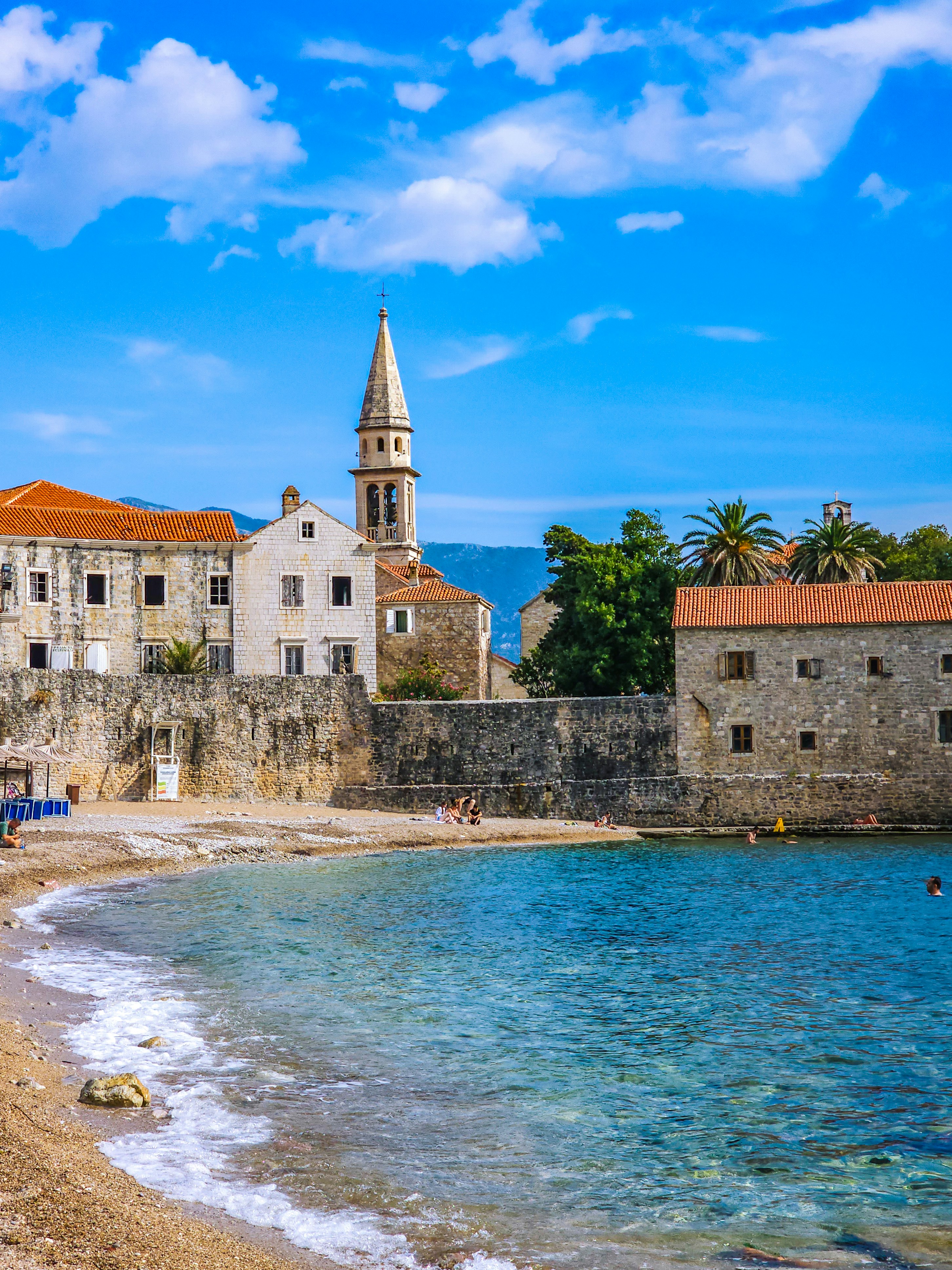 Historic coastal buildings with a bell tower overlooking a serene beach and clear waters, framed by palm trees and a vibrant sky.