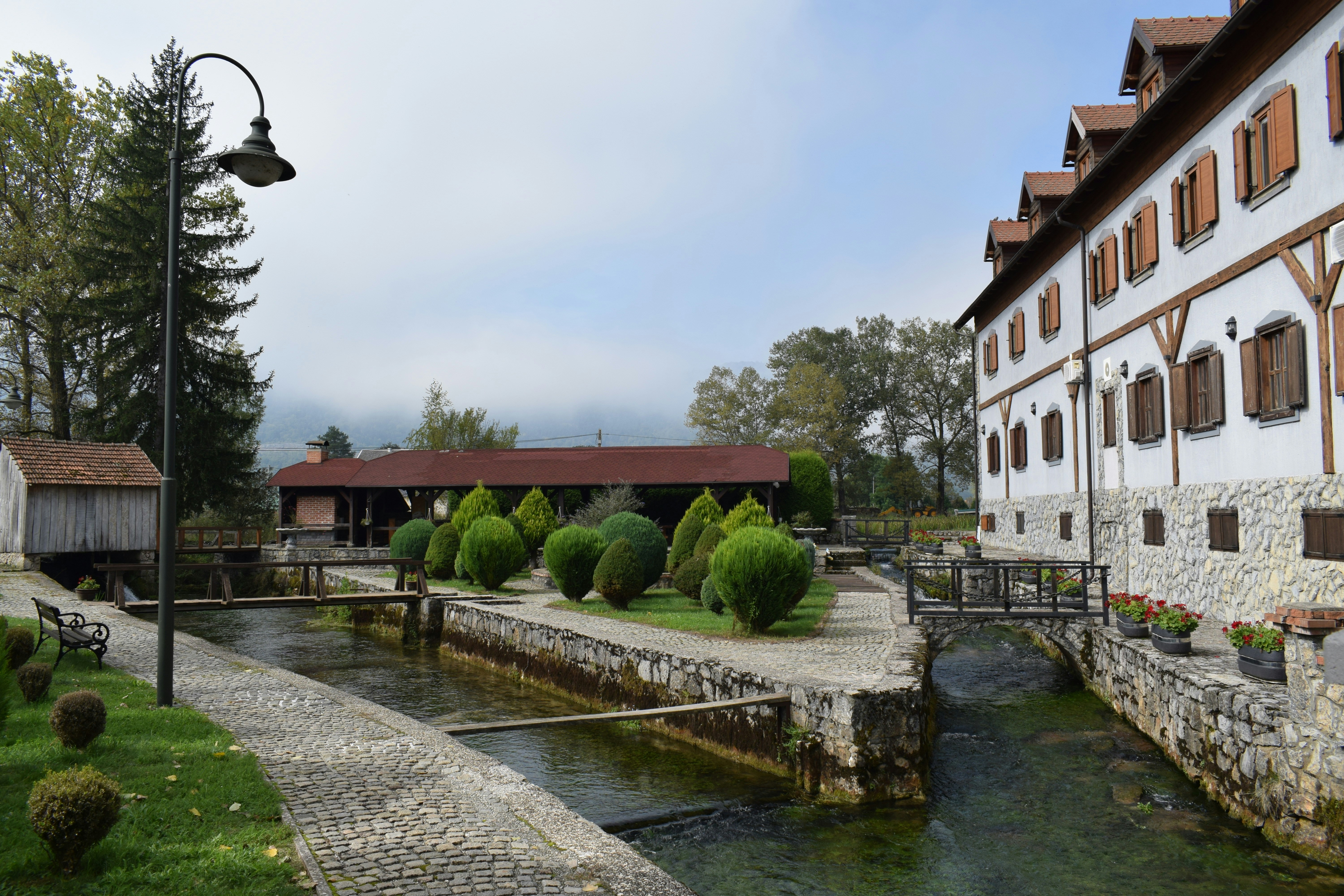 Rustic hotel beside a serene stone-lined river and lush greenery under a partly cloudy sky.