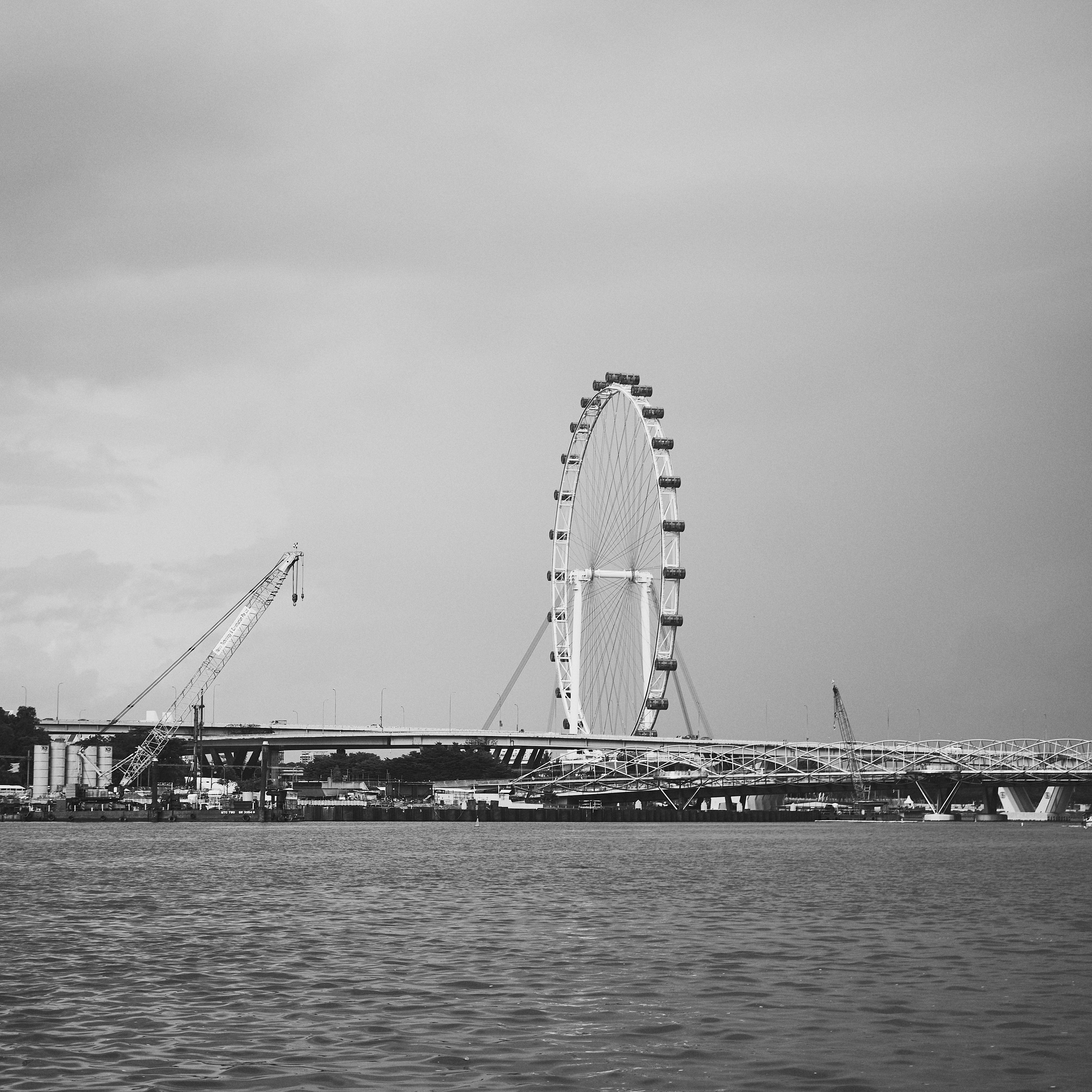 A large Ferris wheel stands against a cloudy sky, framed by construction cranes and a bridge, highlighting the blend of leisure and industry.