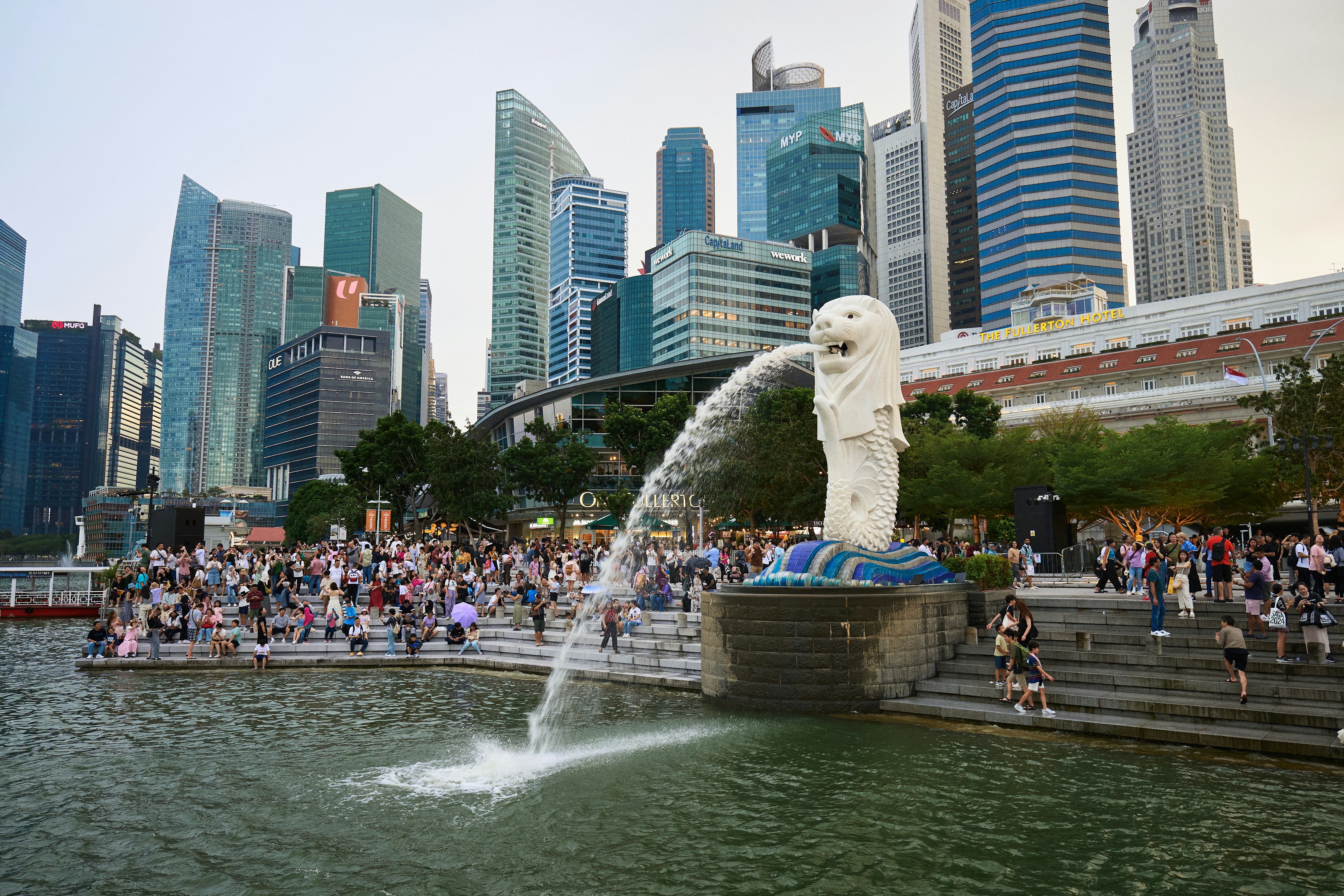 Water jets from the Merlion at Marina Bay as crowds gather on the steps, with Singapore's glass-clad skyline looming behind.