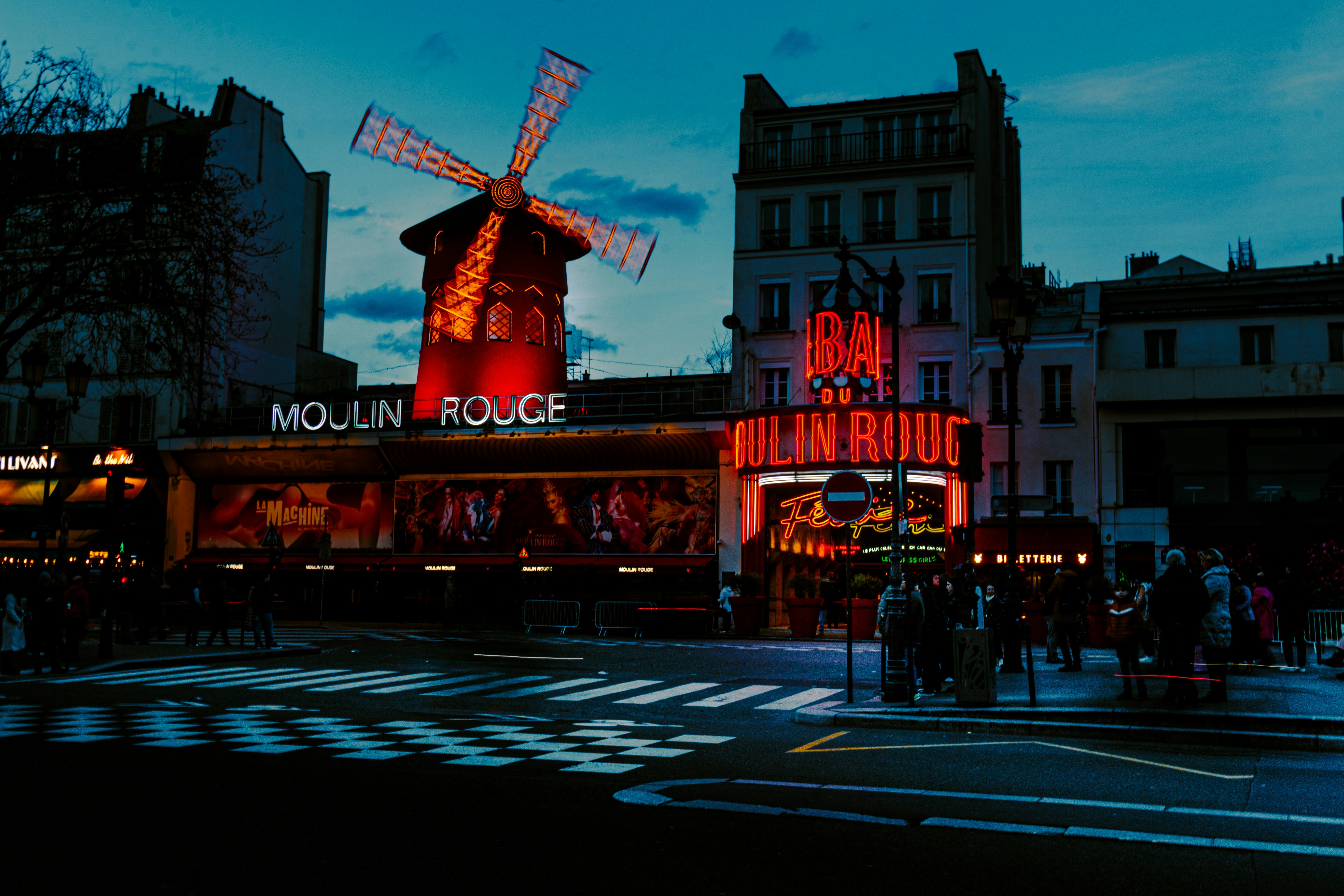 Illuminated windmill atop a vibrant building at dusk, with bustling street in foreground.