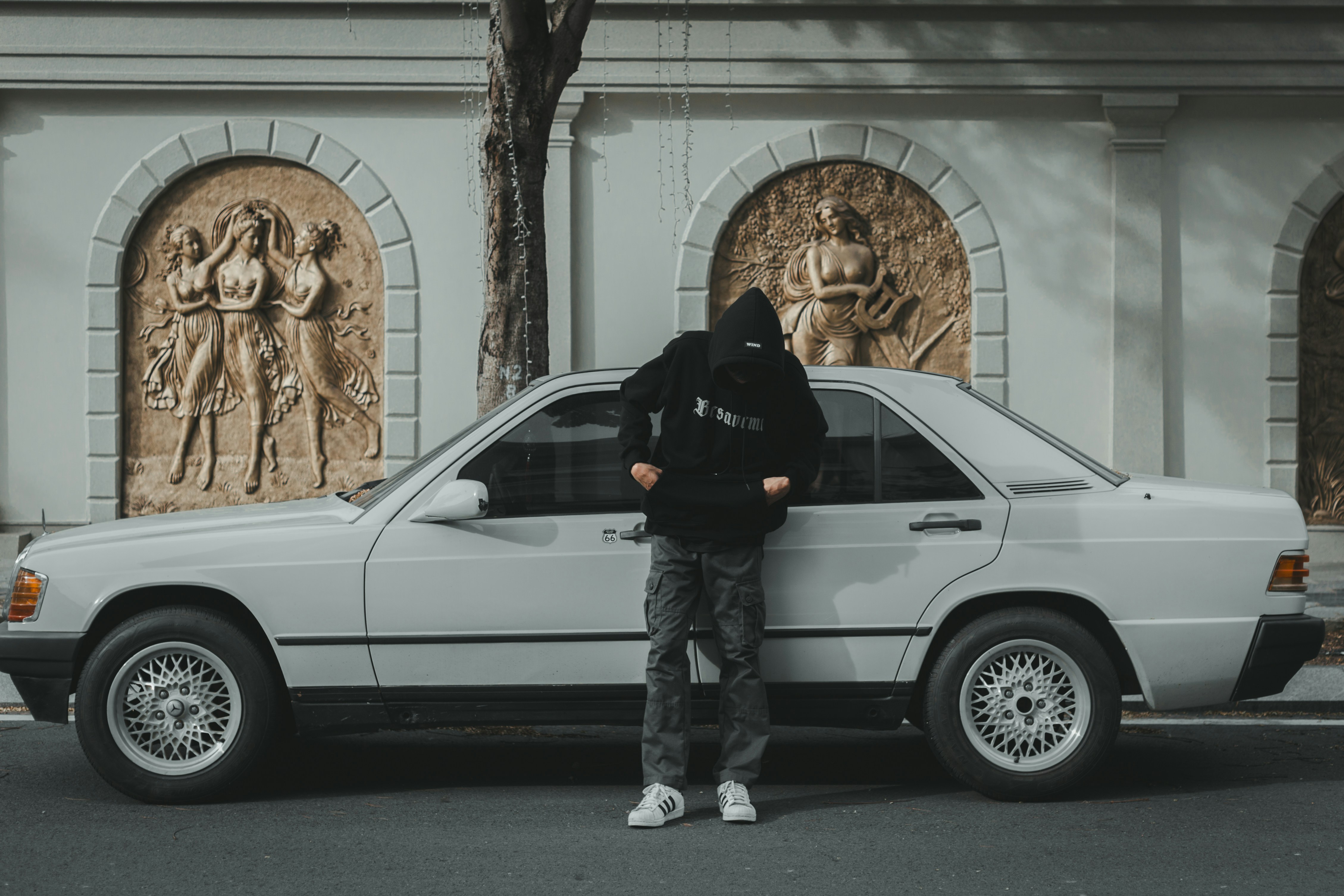 Hooded Stance by a White Classic Sedan Against Bronze Carvings