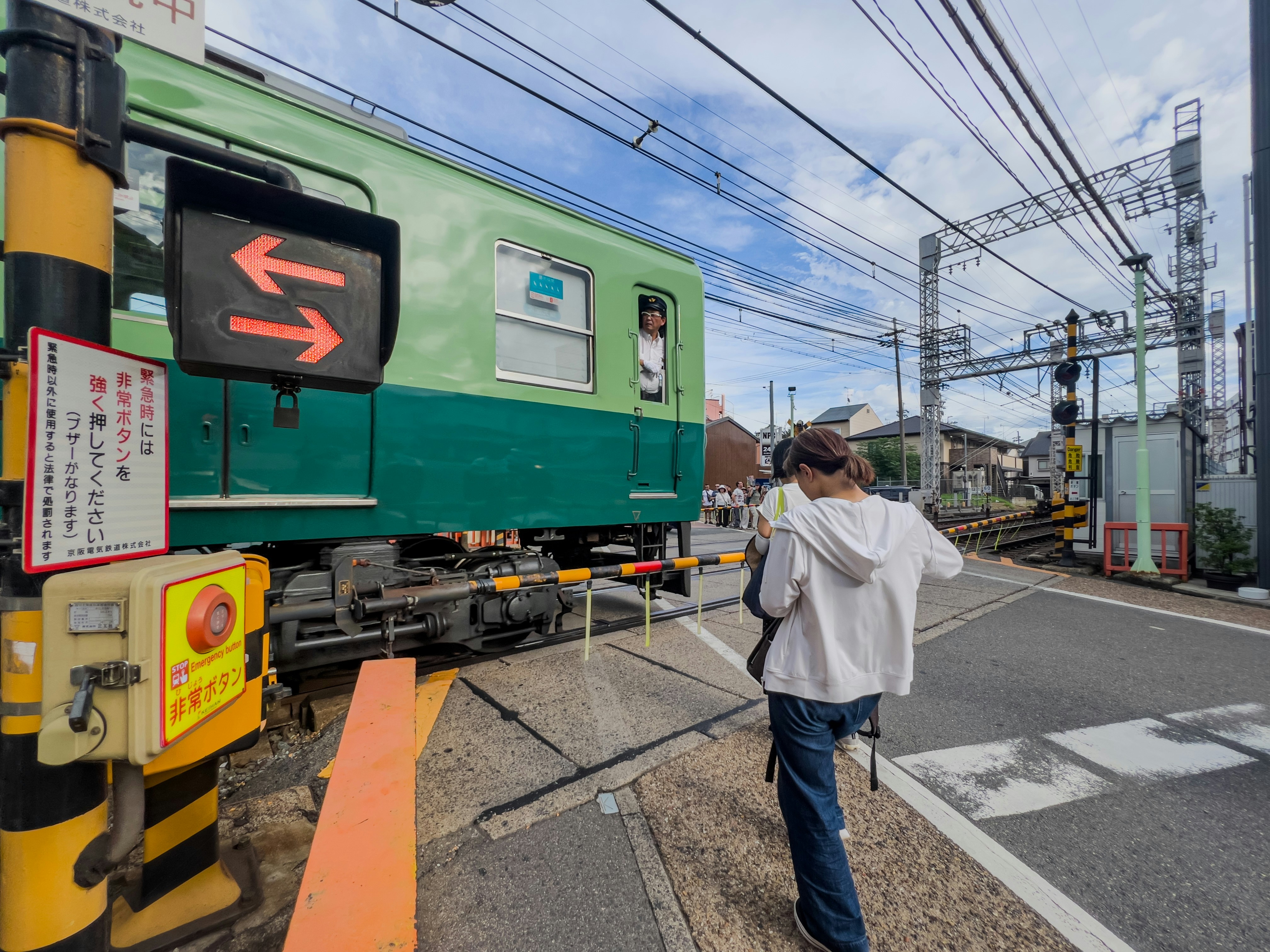 A green train approaches a railway crossing, while a person stands nearby, observing the scene. The image captures the intersection of daily life and public transportation.