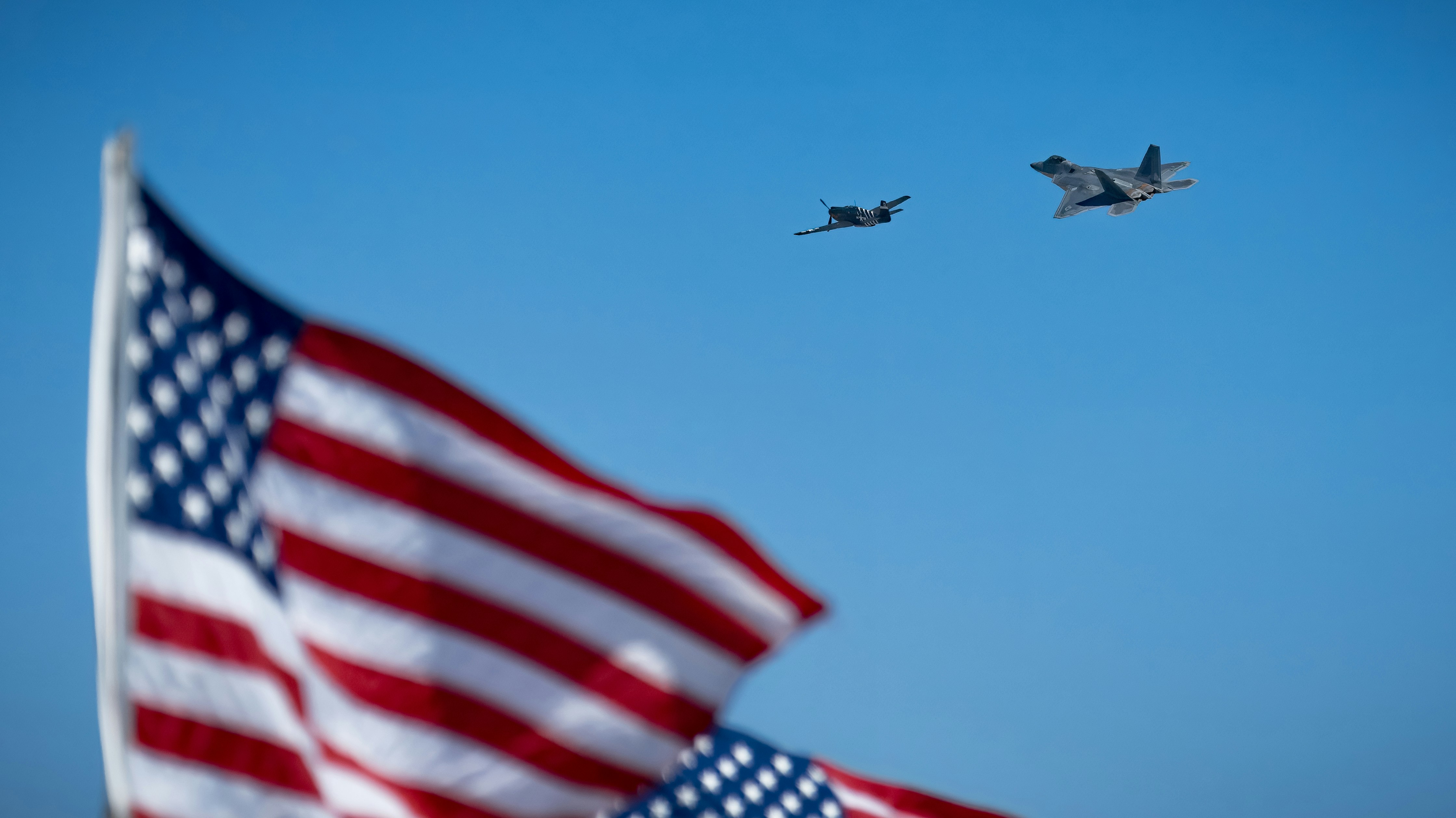 American flag waves in foreground with two military aircraft flying in formation against a clear blue sky.