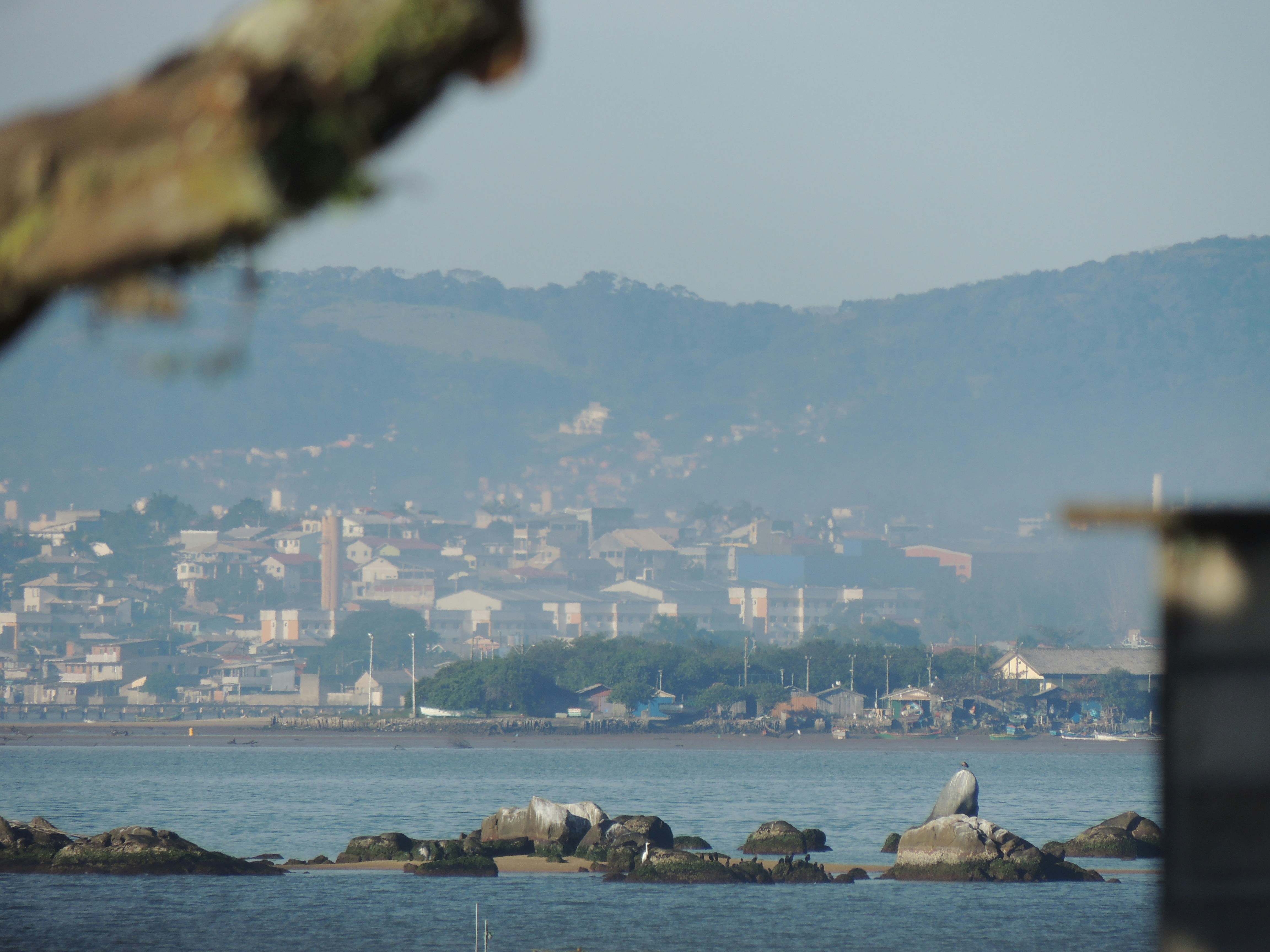 Distant cityscape viewed across a calm bay with a mountainous backdrop and scattered rocks in the foreground.