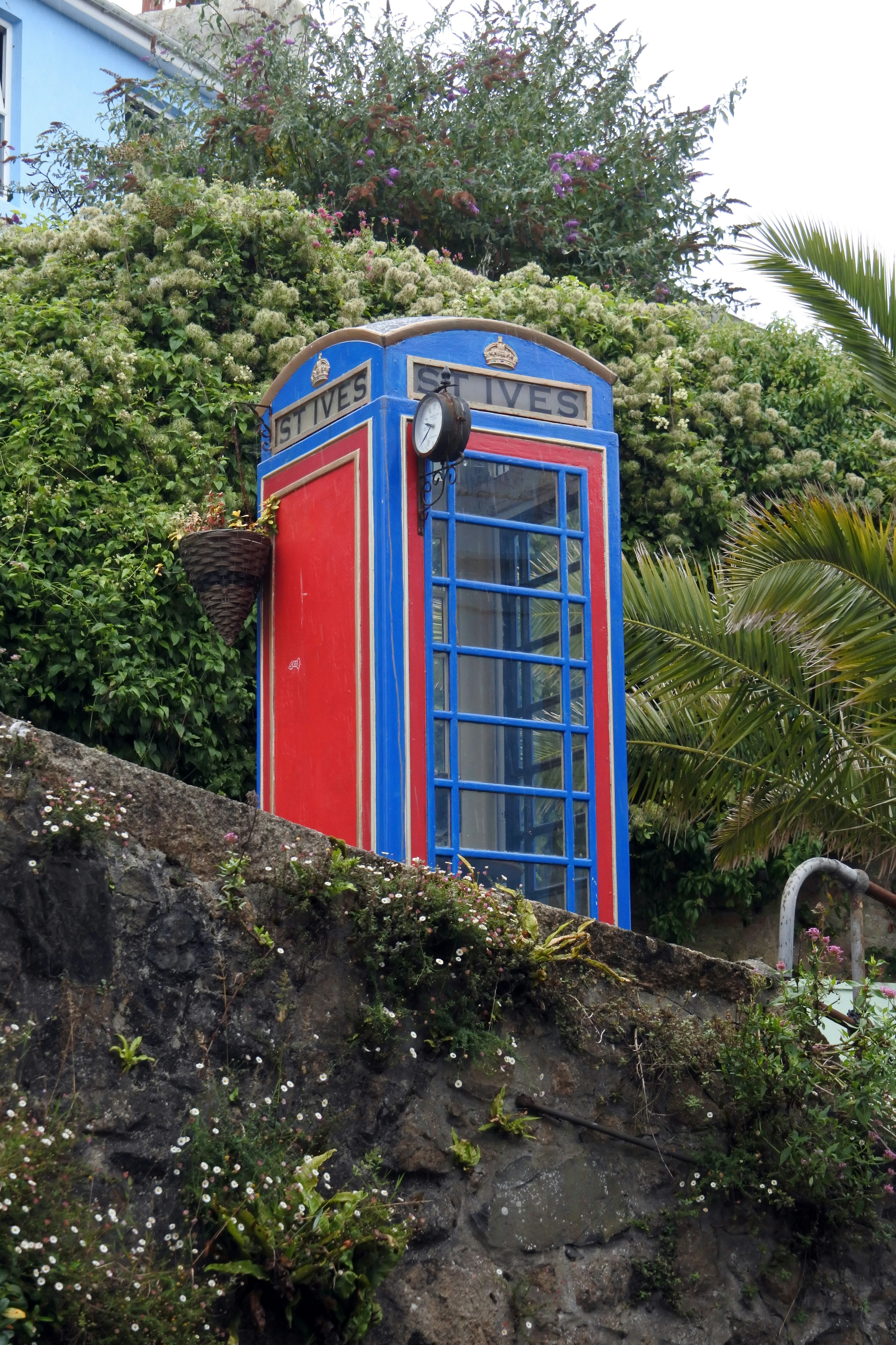 A bright red telephone booth with blue trim sits on a rocky terrace surrounded by lush greenery and flowering plants.
