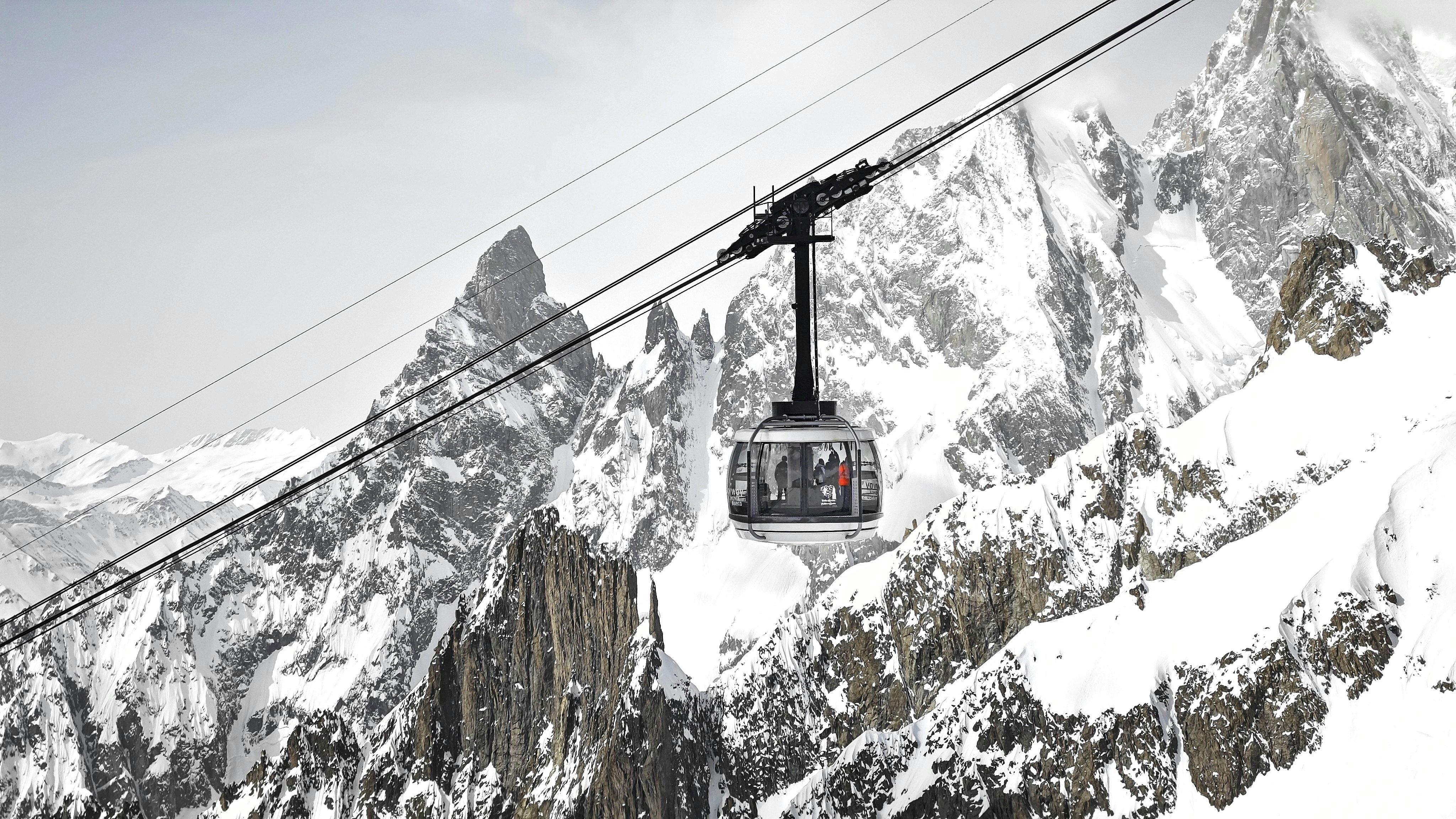 Cable car glides between jagged, snow-clad peaks over a rugged alpine landscape. The image emphasizes altitude and the austere beauty of winter mountains.