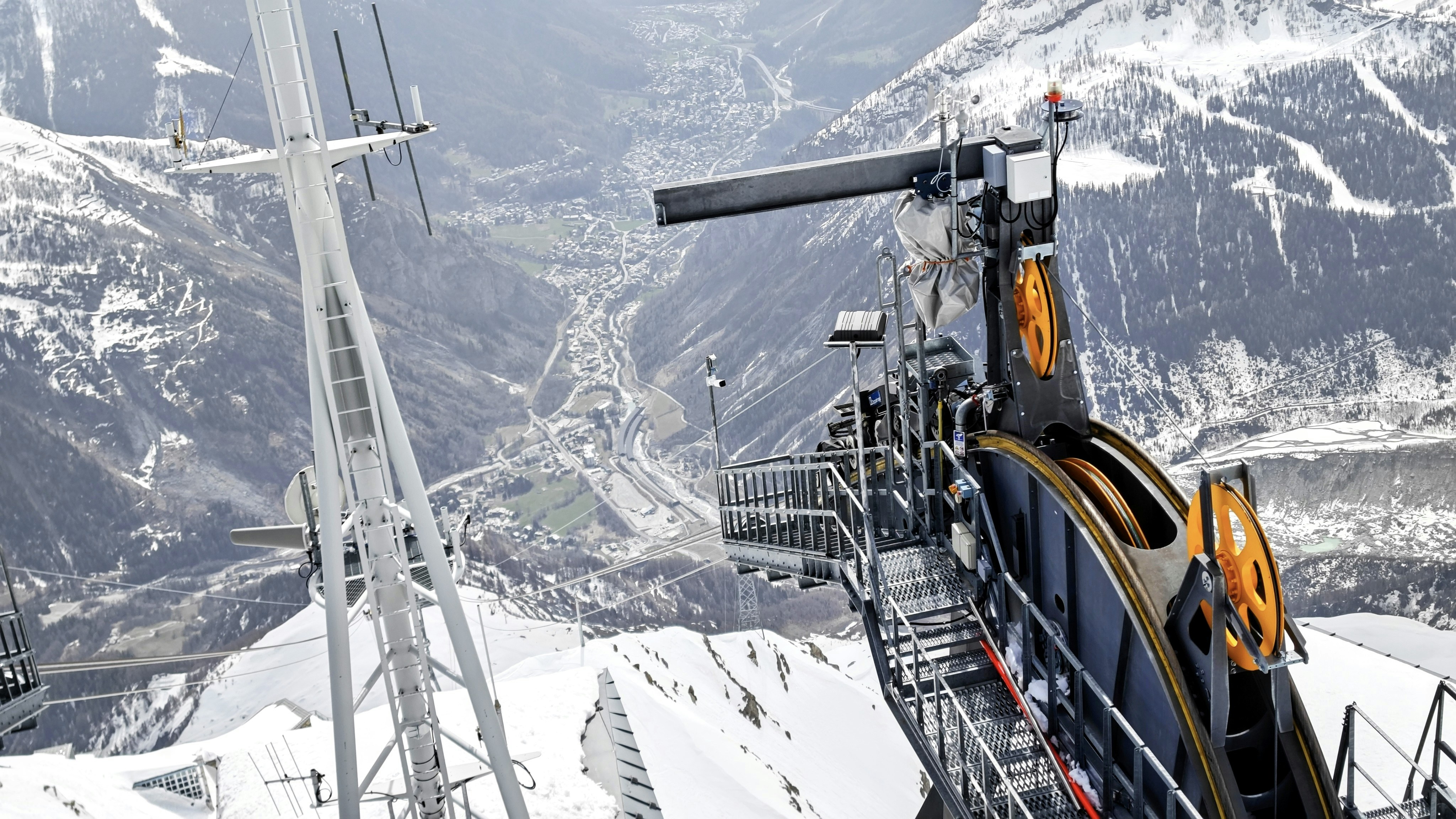Snowy alpine landscape with a cable car lift, tall towers spanning the slope and orange wheels visible on the right.