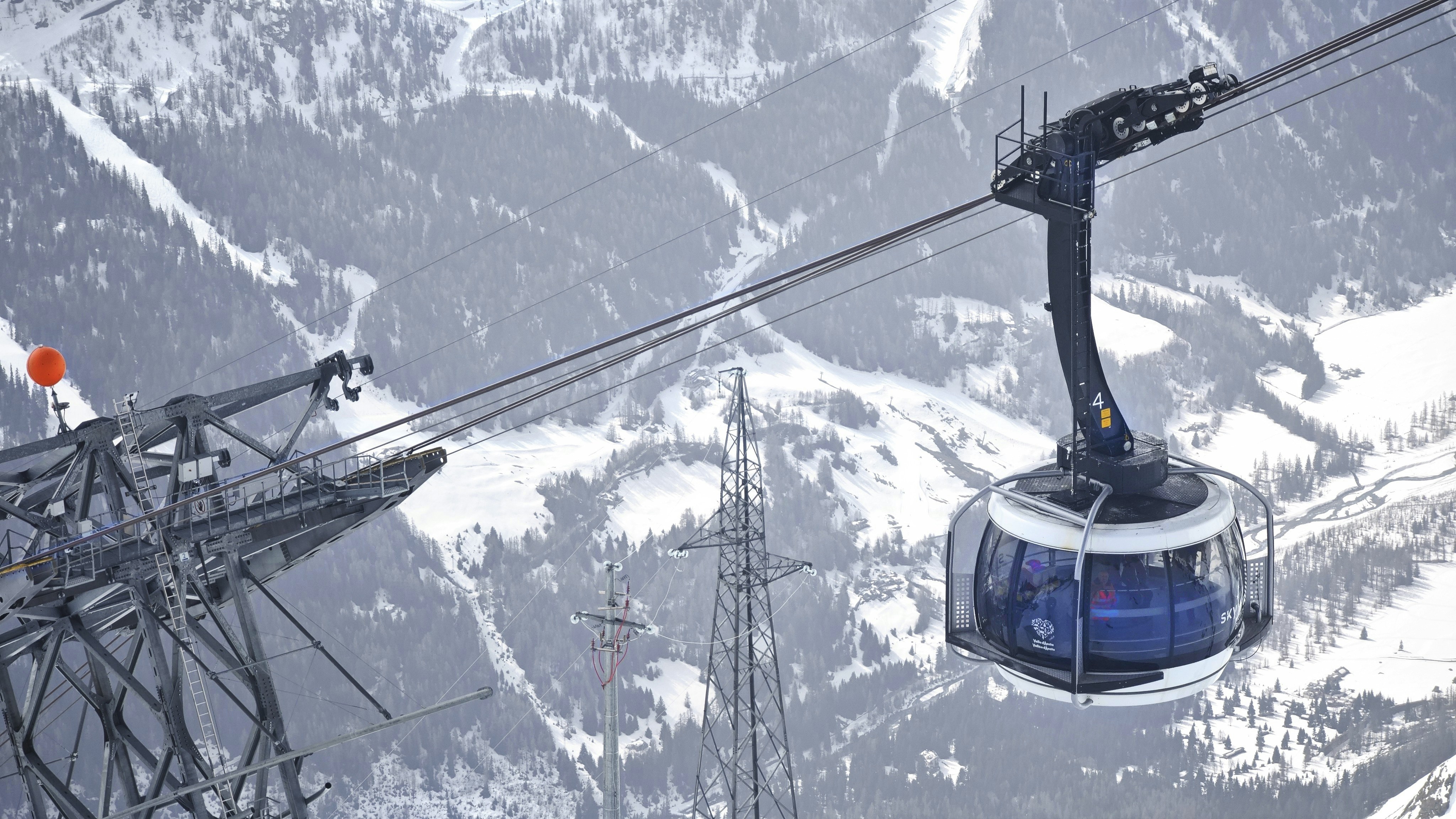 Photograph of a cable car gliding above snow-covered alpine slopes, with towers and cables spanning a frosty valley.