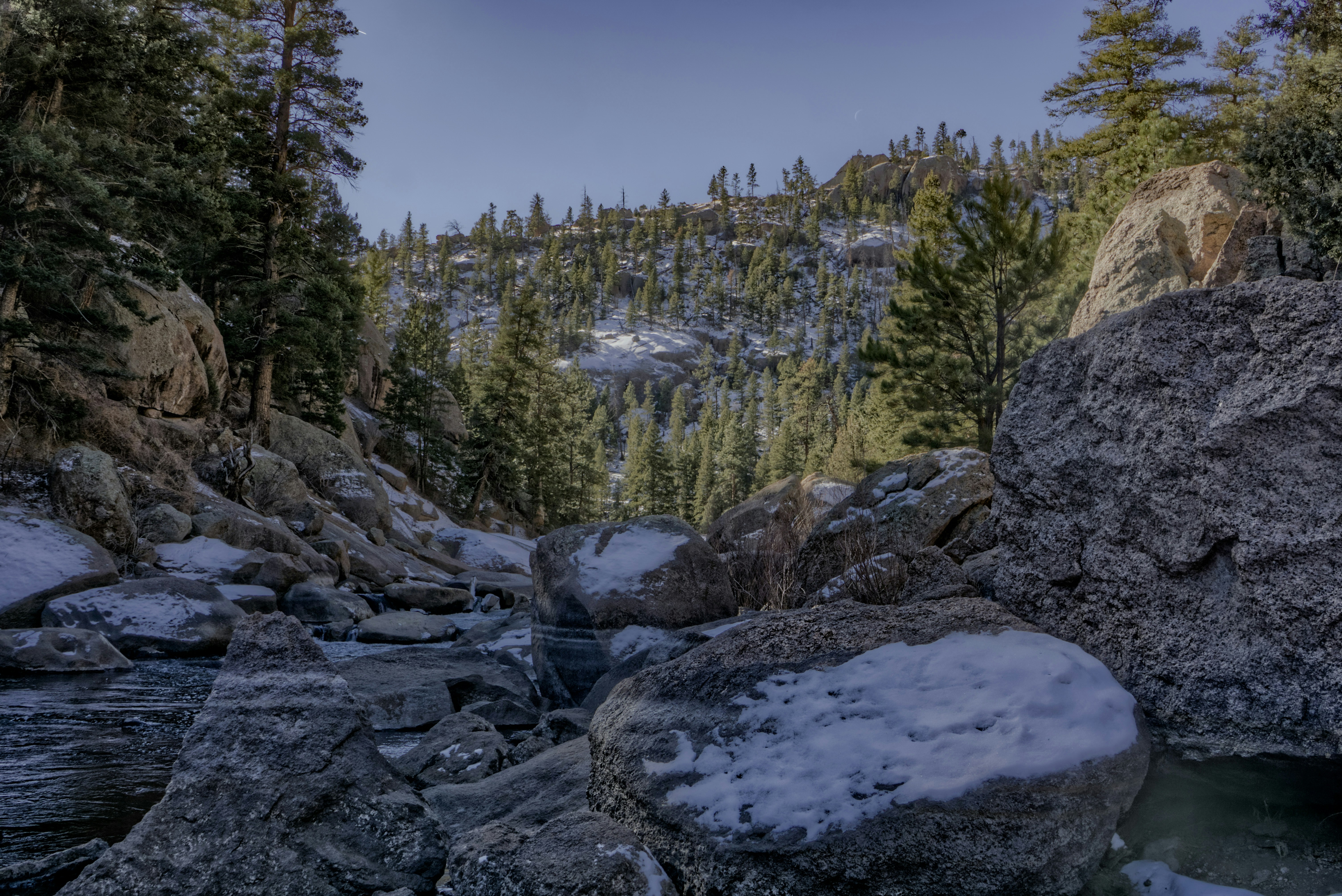 Snow-dusted rocks and flowing river framed by evergreen trees under a clear sky.