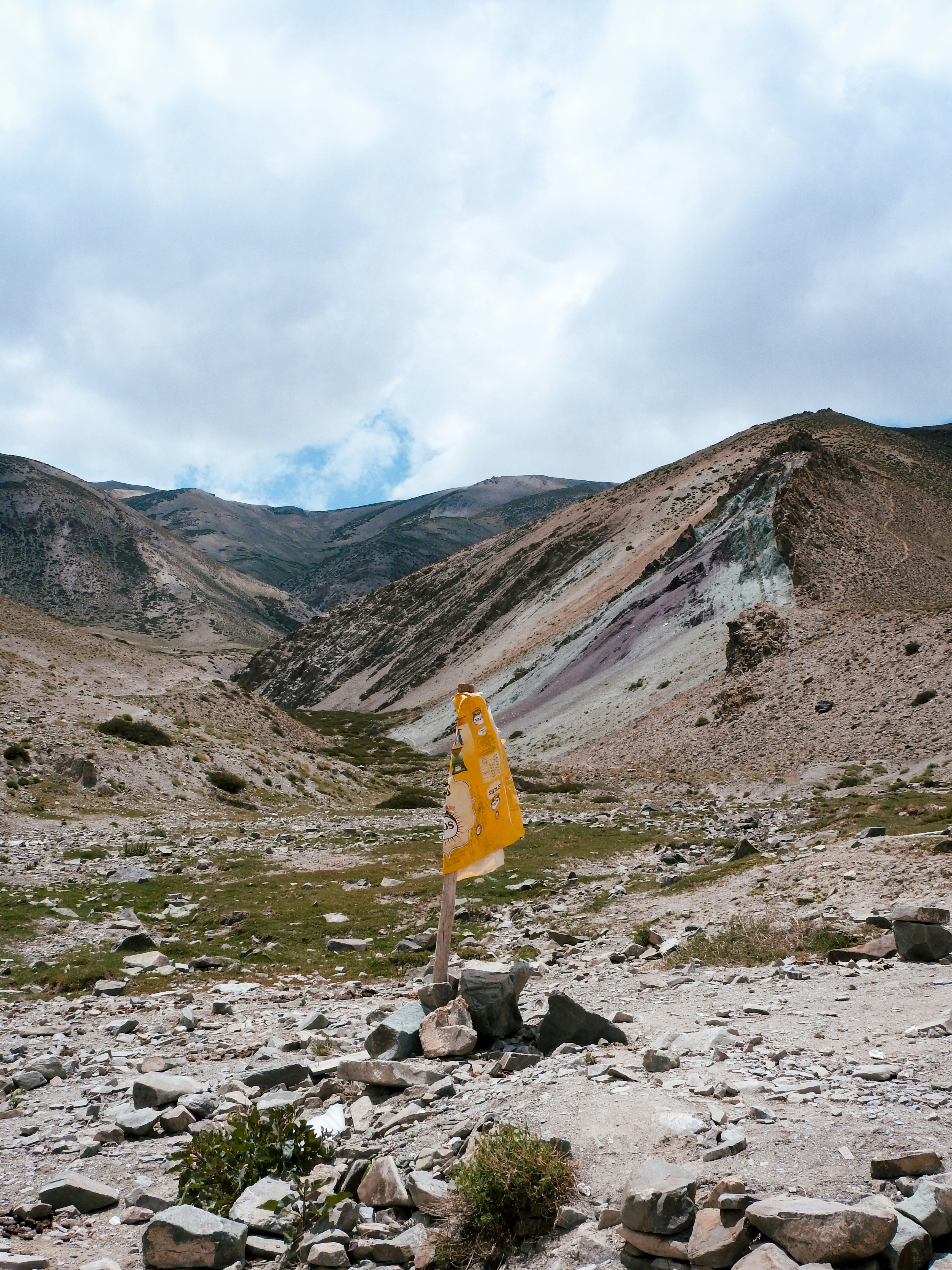 A lone yellow flag flutters atop a weathered post amid a pebble-strewn basin, with eroded hills and a cloudy sky in the background.