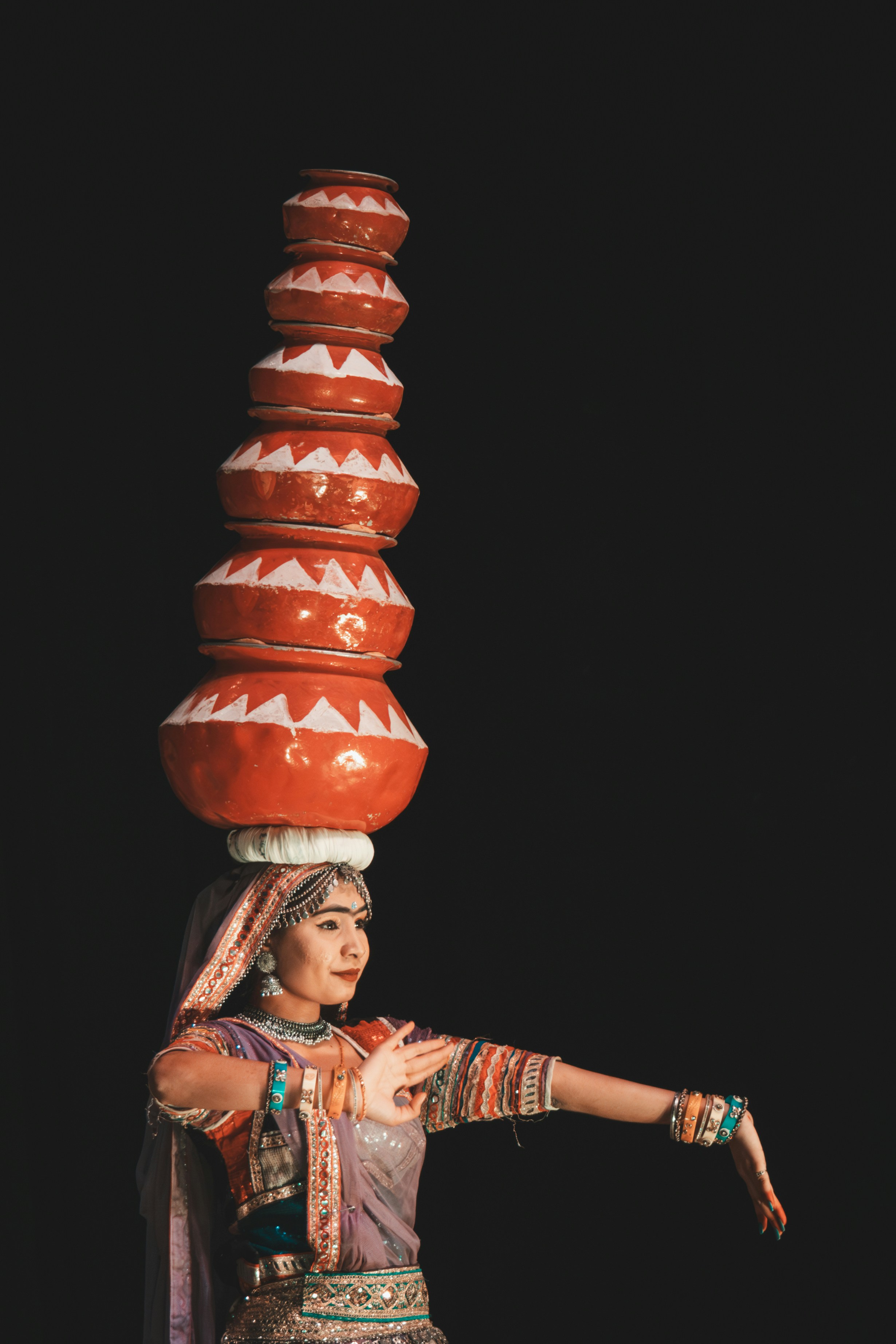 A dancer gracefully balancing intricately painted clay pots atop her head, showcasing traditional attire and expressive hand gestures against a dark backdrop.