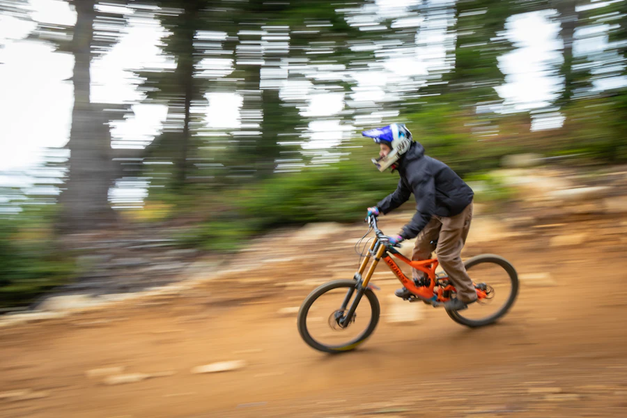 A helmeted mountain biker rides fast along a dusty forest trail, with motion-blurred trees streaking across the background.