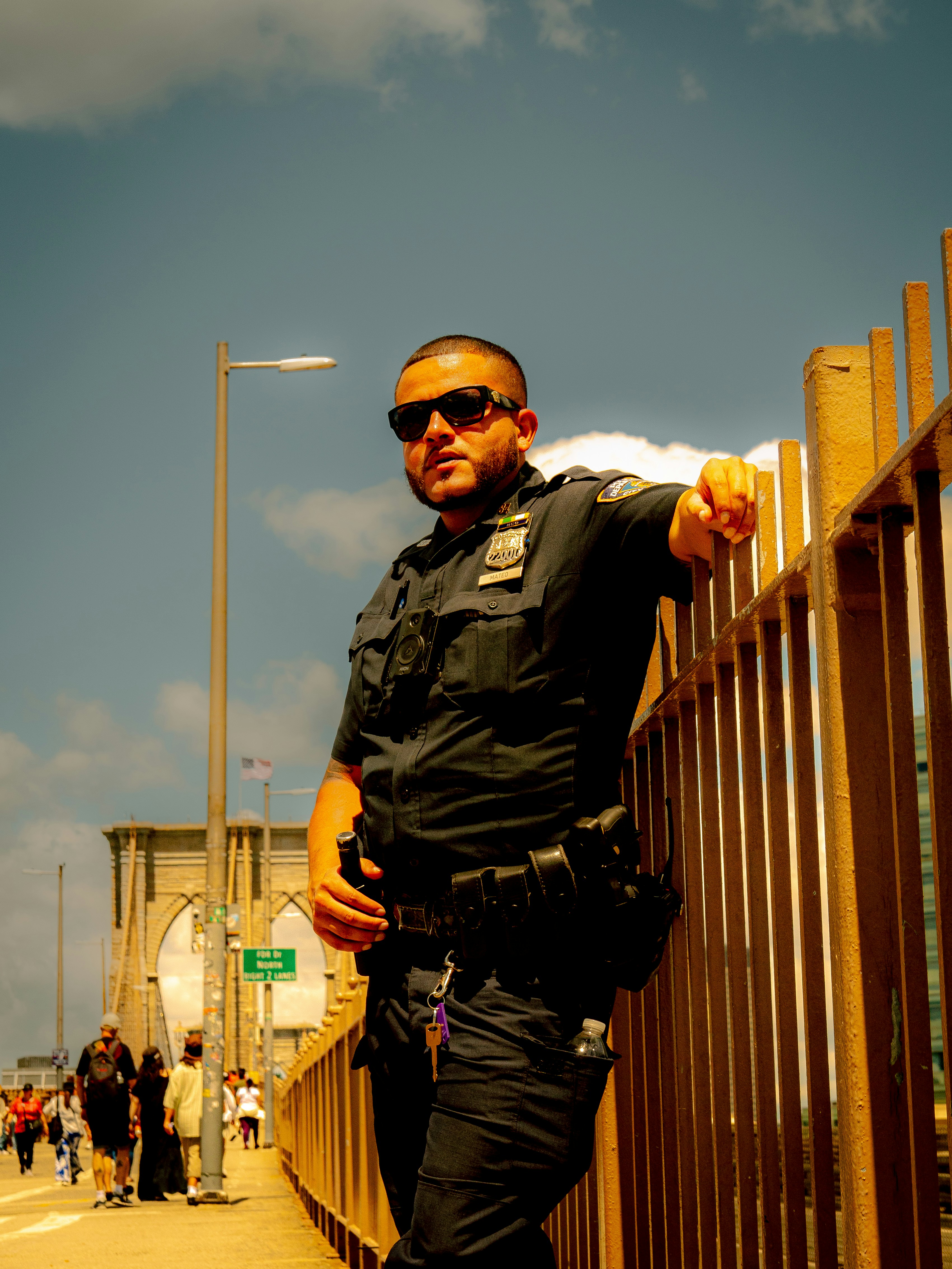 Police officer standing confidently by a railing on the Brooklyn Bridge, with pedestrians in the background. The scene captures urban life and the role of law enforcement.