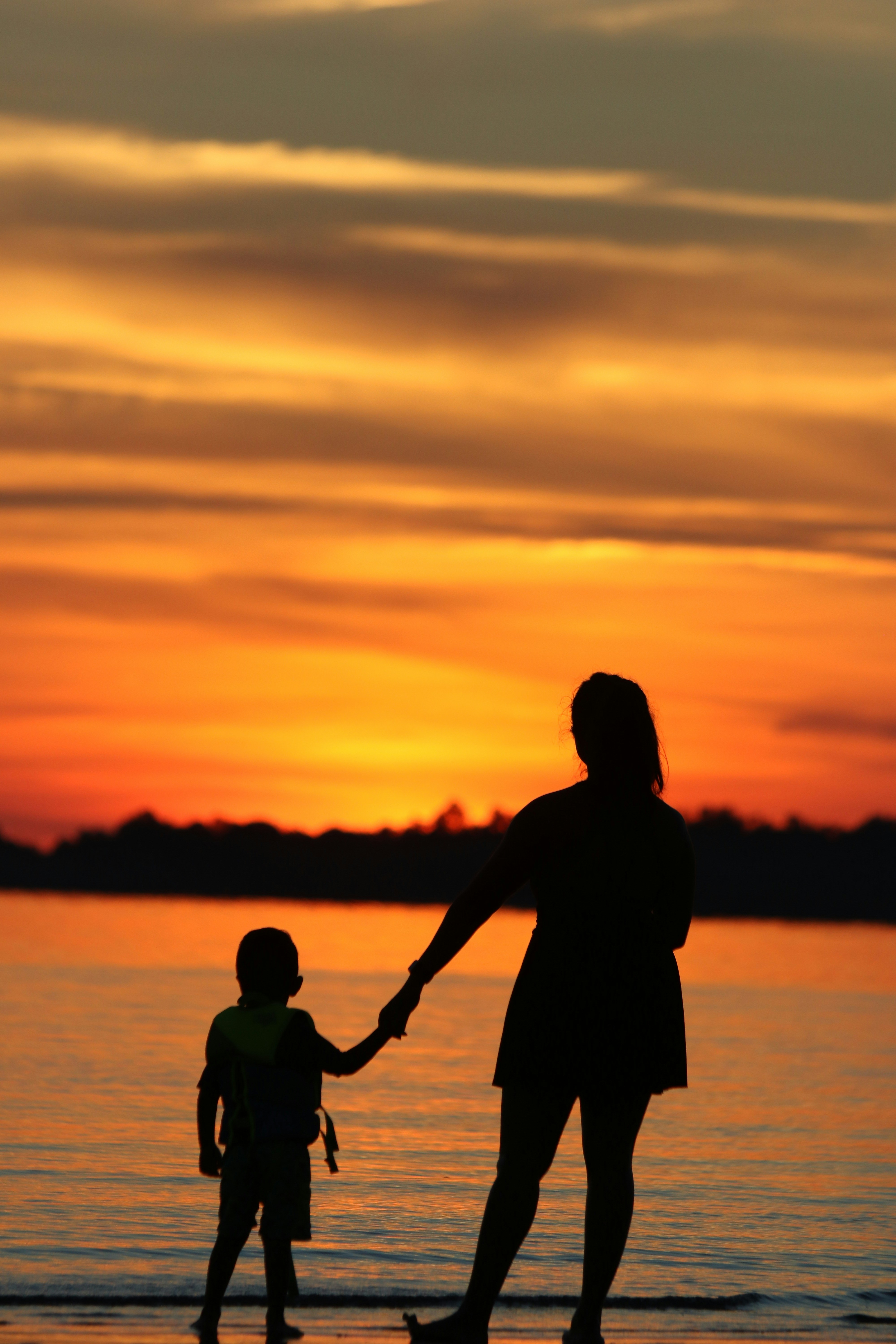 Silhouetted figures of an adult and child holding hands against a vibrant sunset over calm waters.