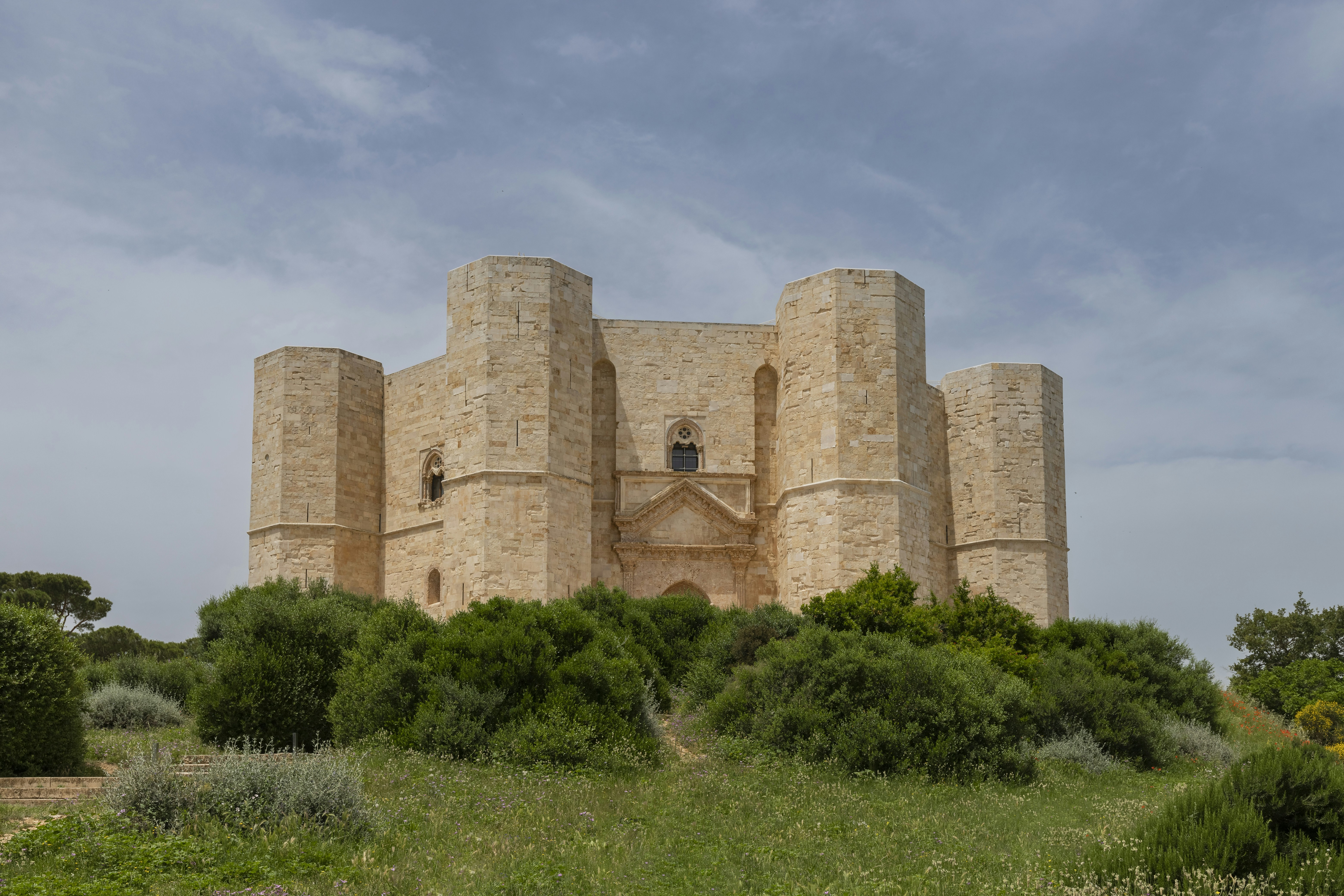 Stone castle with central tower surrounded by lush greenery under a cloudy sky.