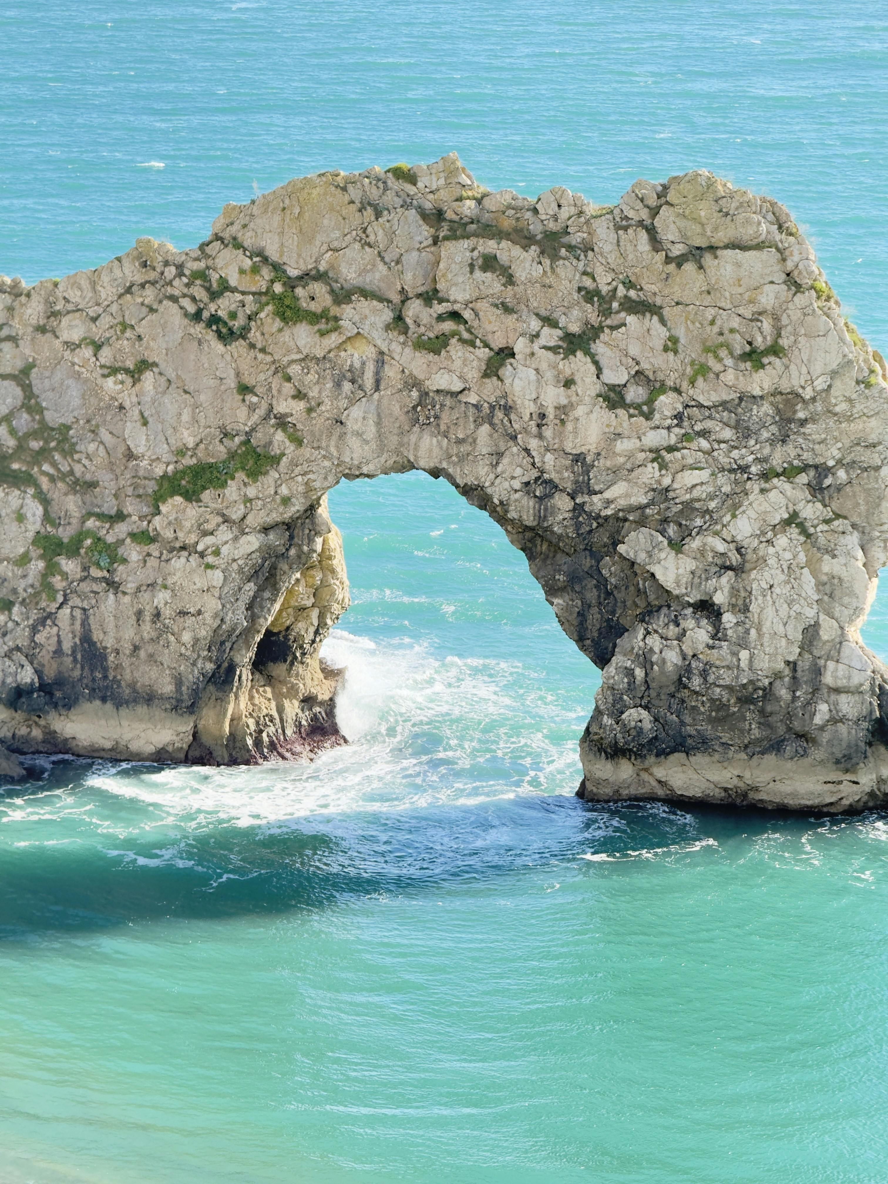 Natural rock formation arching over turquoise waters, with waves crashing against its base.