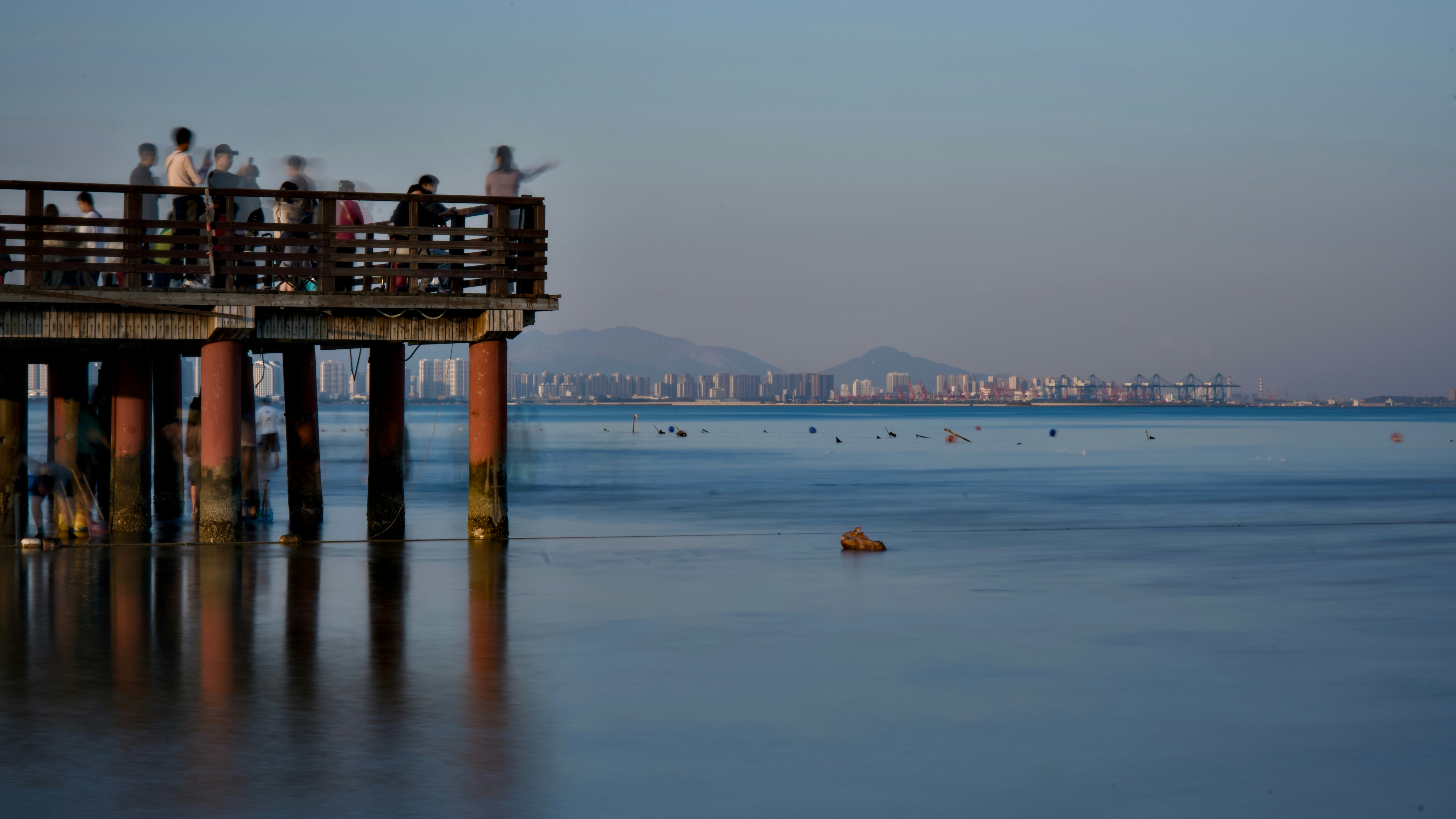 People standing on a pier overlooking a calm body of water with distant city skyline at dusk.