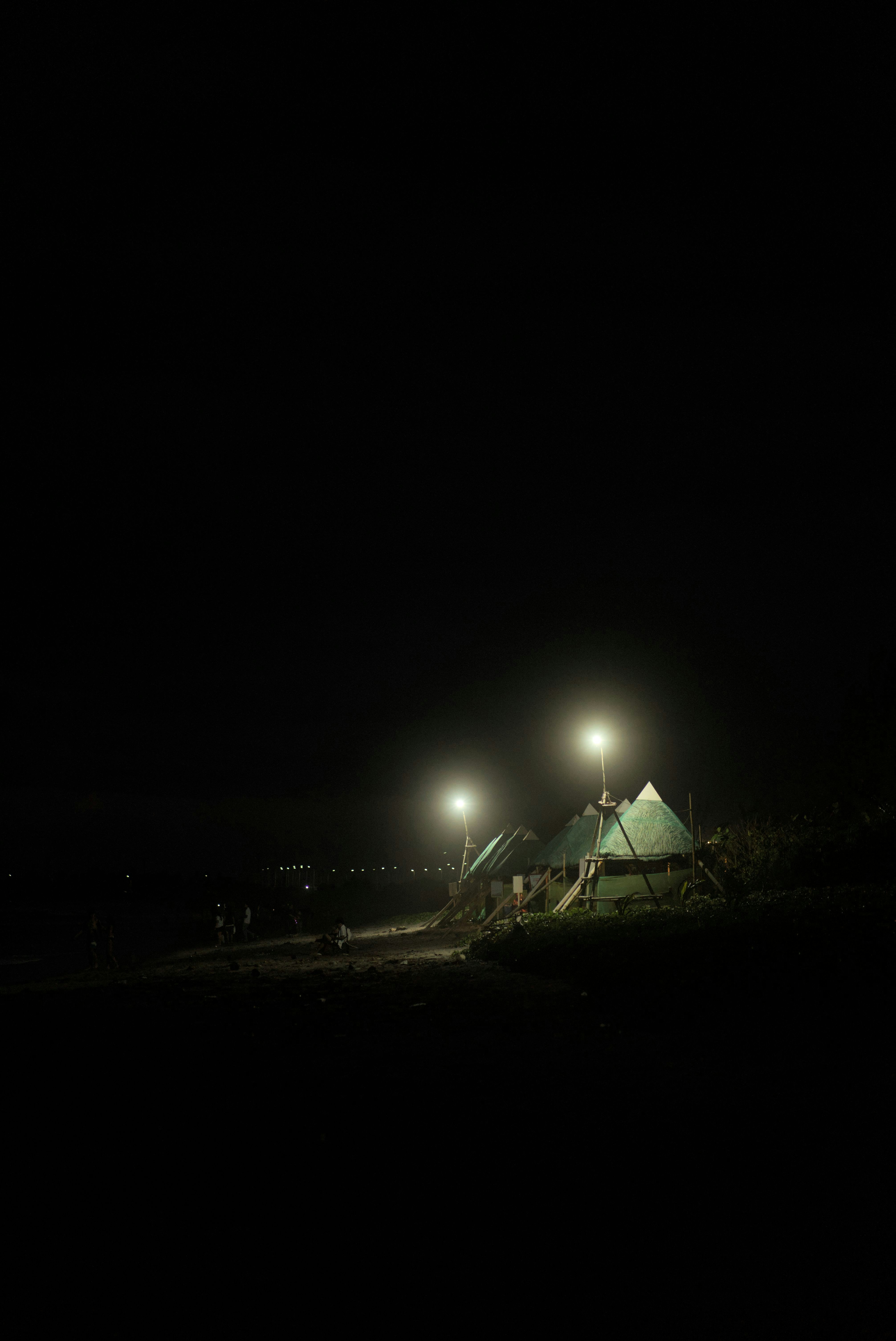 A dark, nighttime scene with two brightly lit huts on a beach. The huts have a conical roof and are illuminated by spotlights.