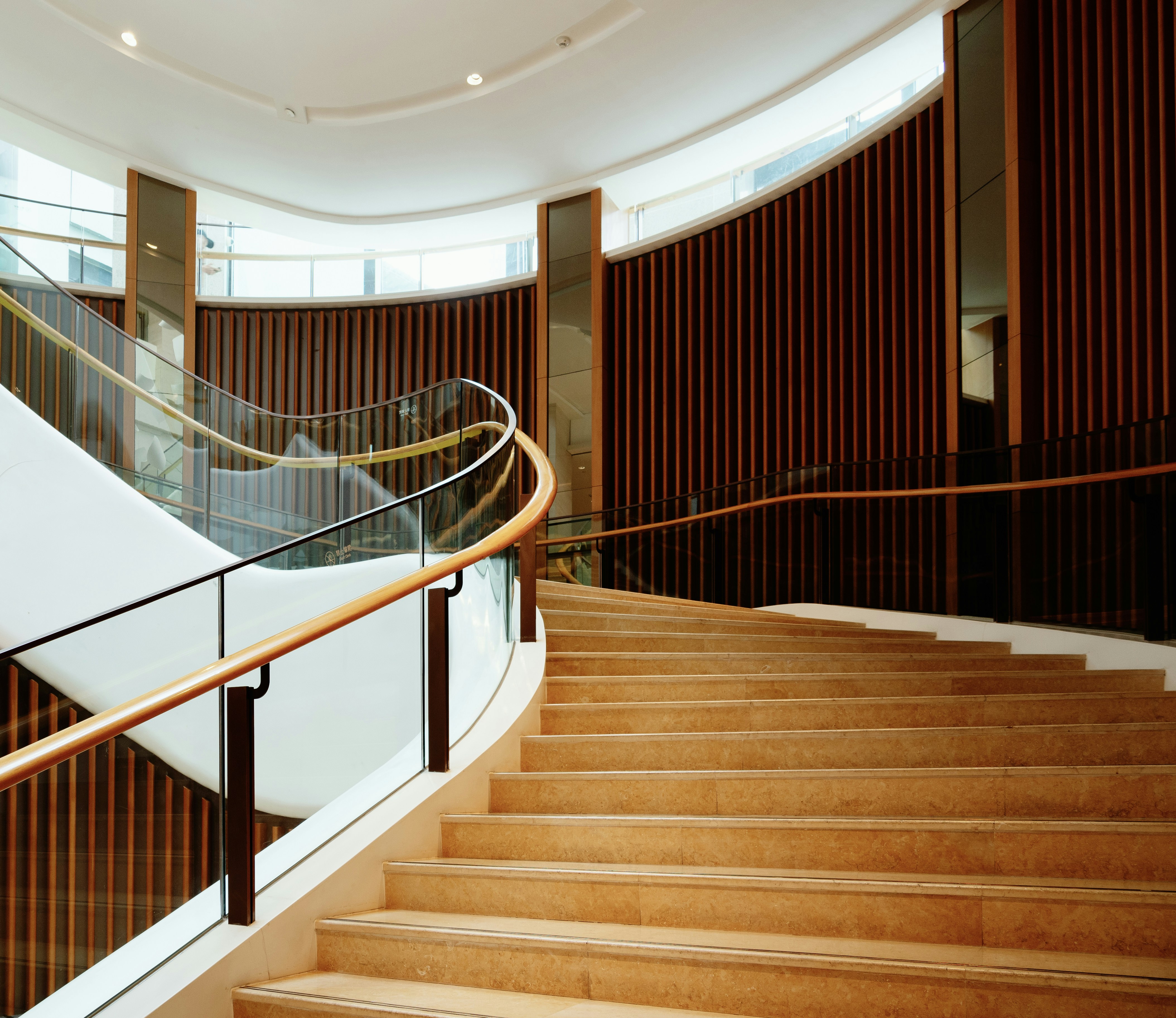 Curved glass railing traces a sweeping maple-wood staircase inside a modern atrium, highlighting architectural rhythm. Soft lighting emphasizes glossy surfaces and the space's refined, quiet mood.