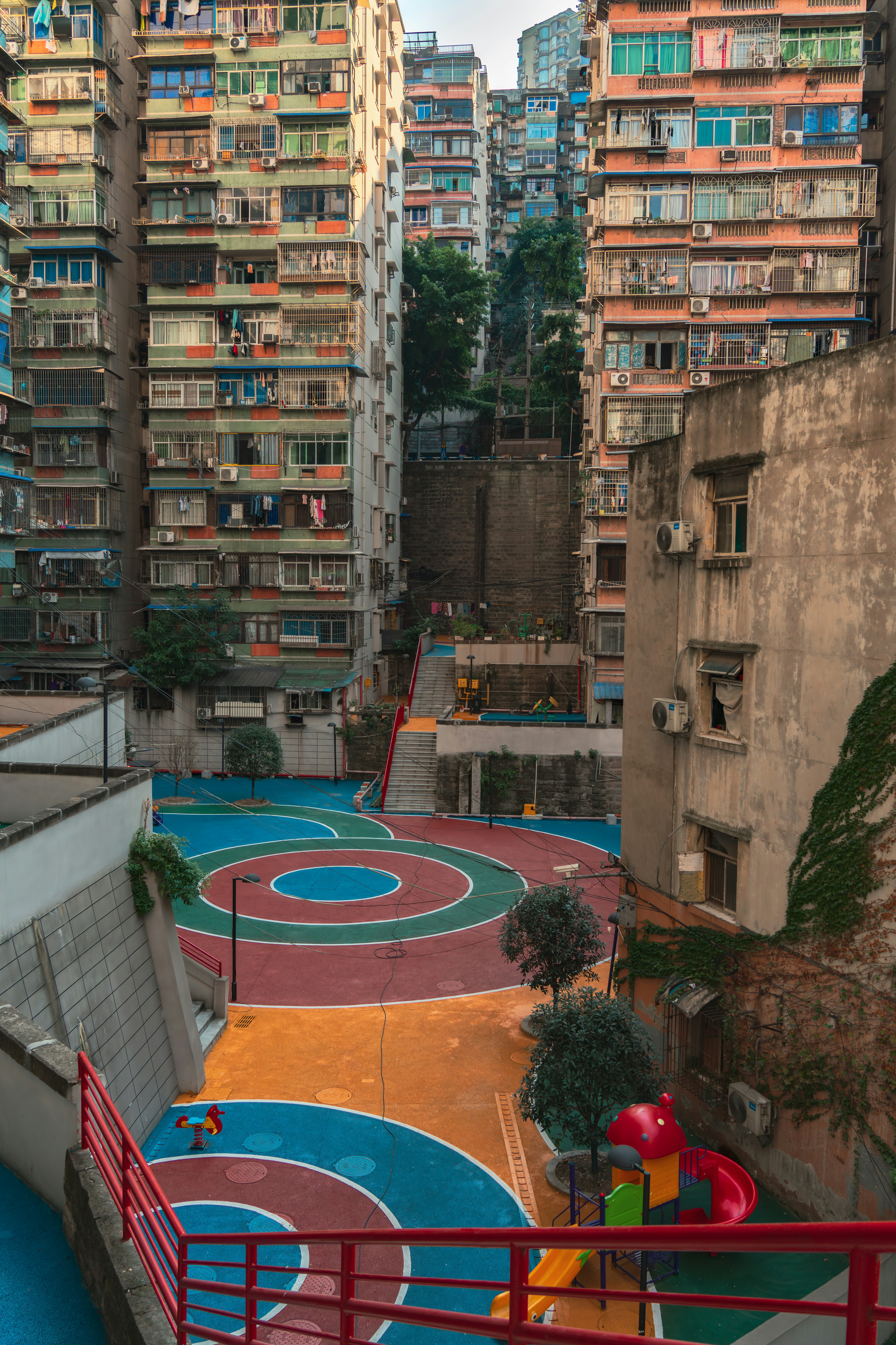 Photograph of a multistory residential block surrounding a vividly colored circular-patterned playground; a red railing frames the foreground. A small play area sits to the right while tall buildings enclose the space.