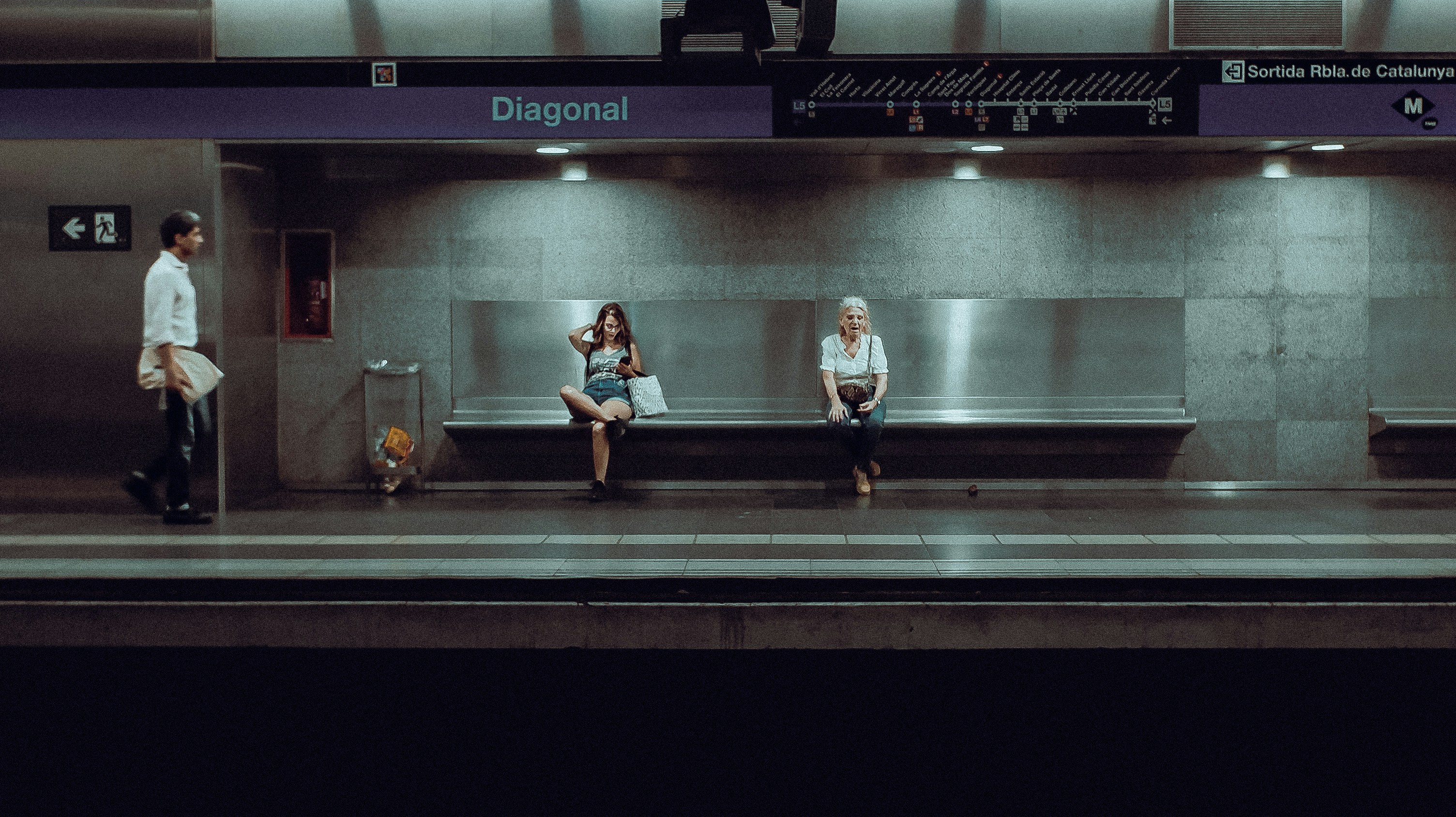 Photograph of three people on a subway platform beneath a 'Diagonal' sign. A man walks by on the left, while a woman sits center-right looking at her phone and another rider sits at the far right.