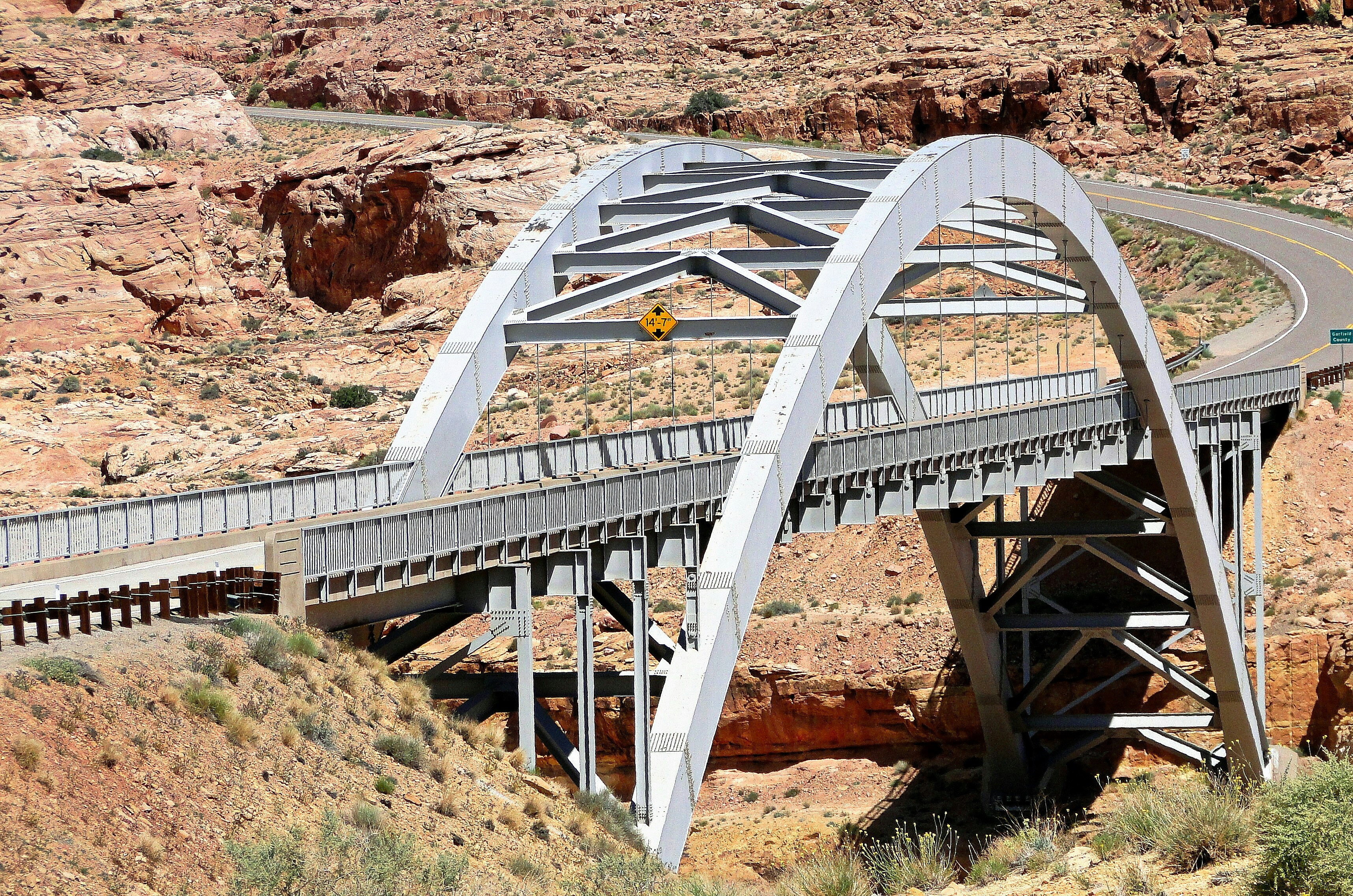 White arch bridge spans a winding road across a desert canyon, with rugged red rock formations in the background.