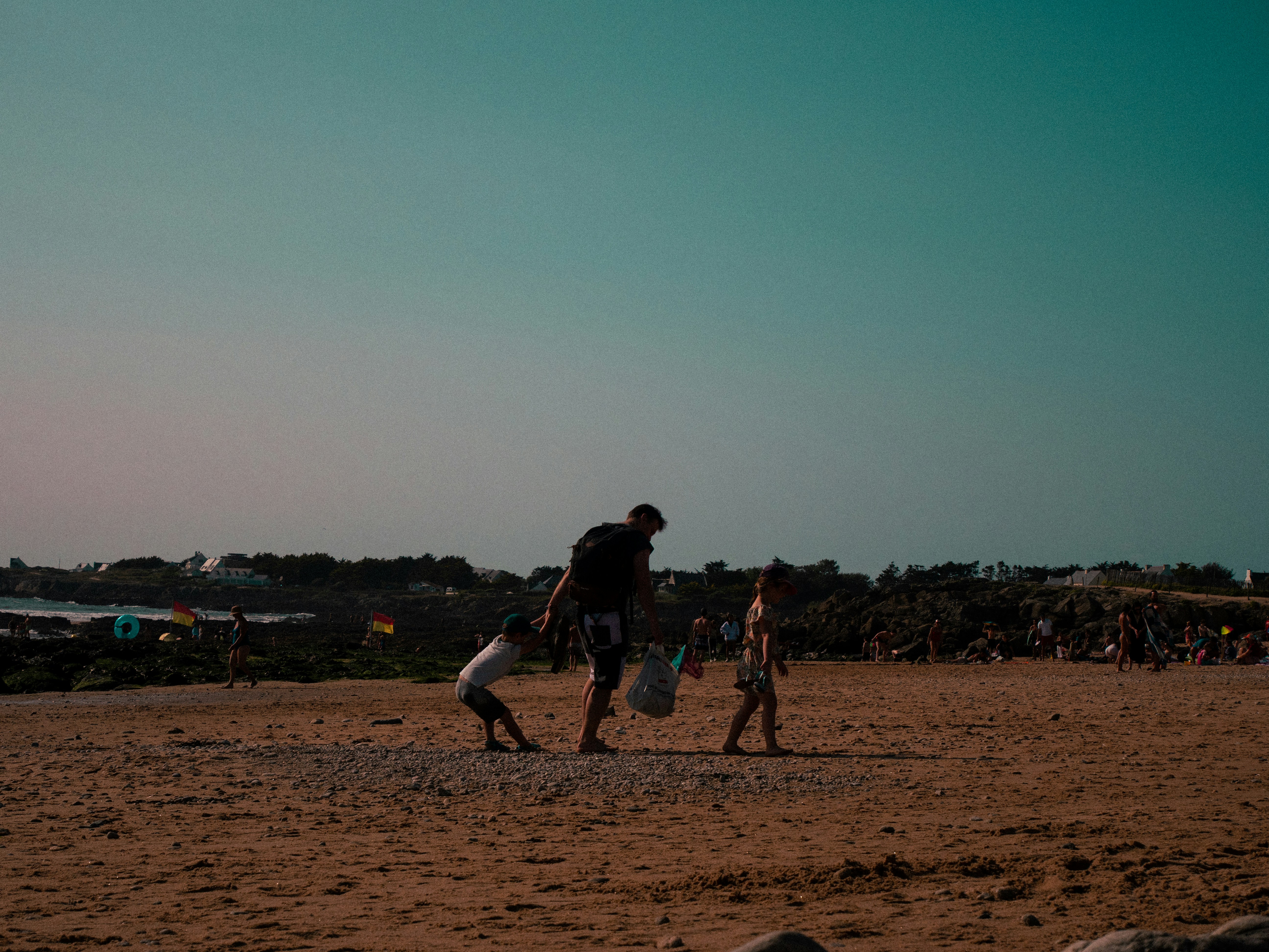 Two figures collecting seashells on a sandy beach, surrounded by a lively atmosphere of sunbathers and distant waves.