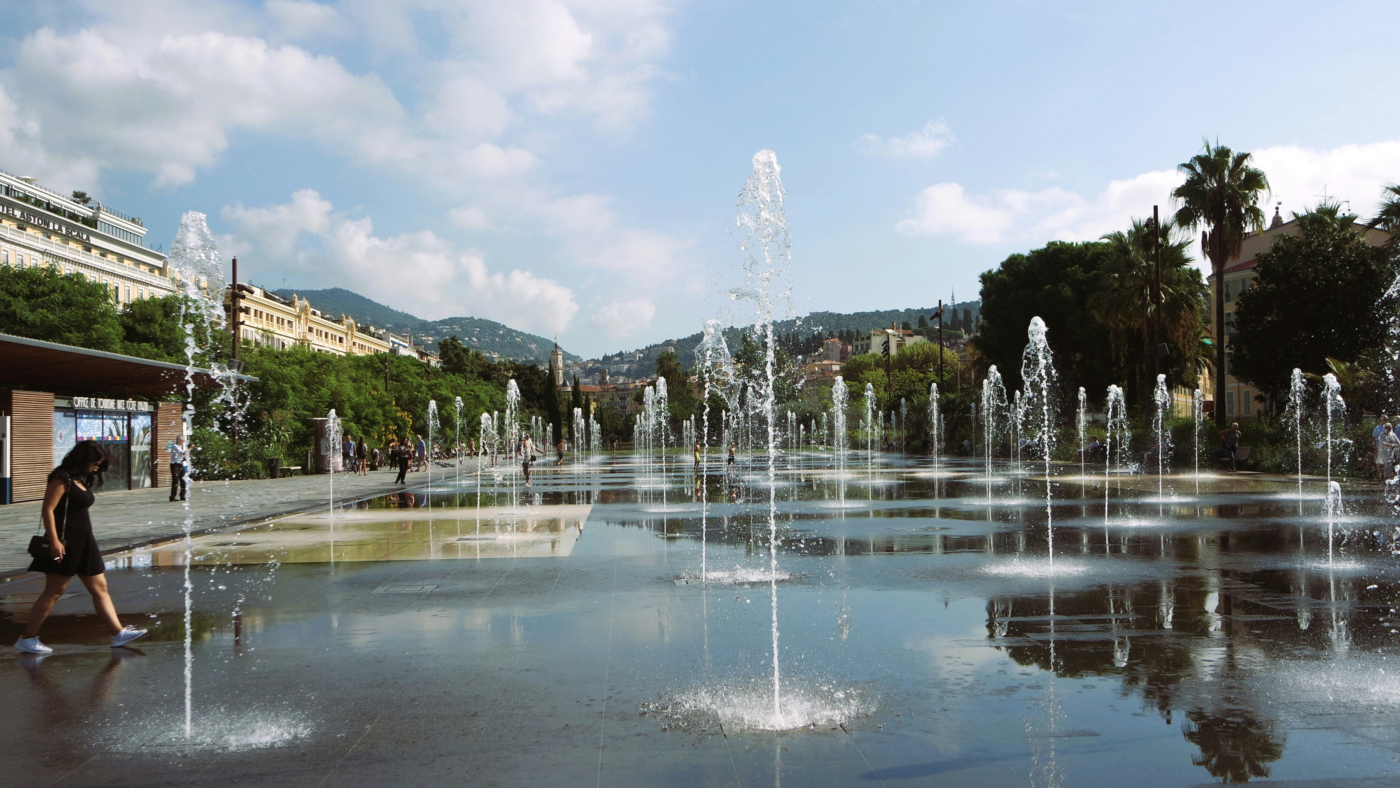 Sunlit city square features multiple water jets shooting from a reflective pool, creating spray and mirrored pavement. A pedestrian walks along the edge as trees and architectural landmarks frame the scene.