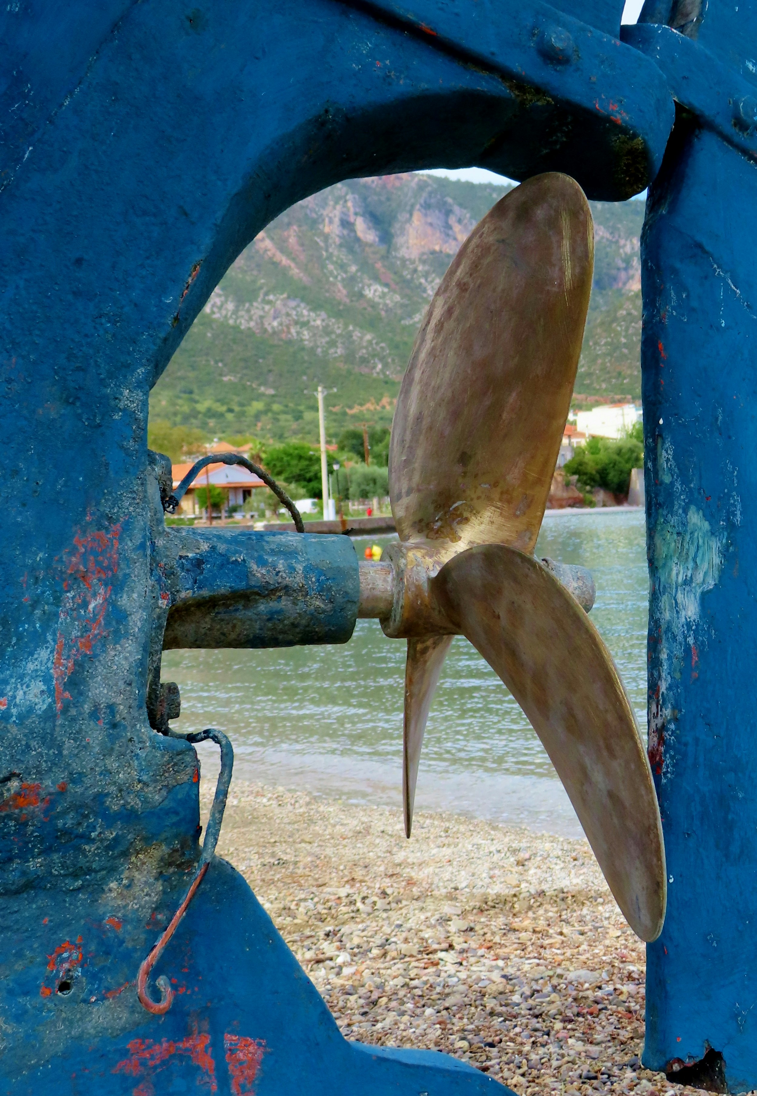 Weathered blue boat frame encloses a brass propeller against a lakeside scene. Distant mountains and a small village line the shore.