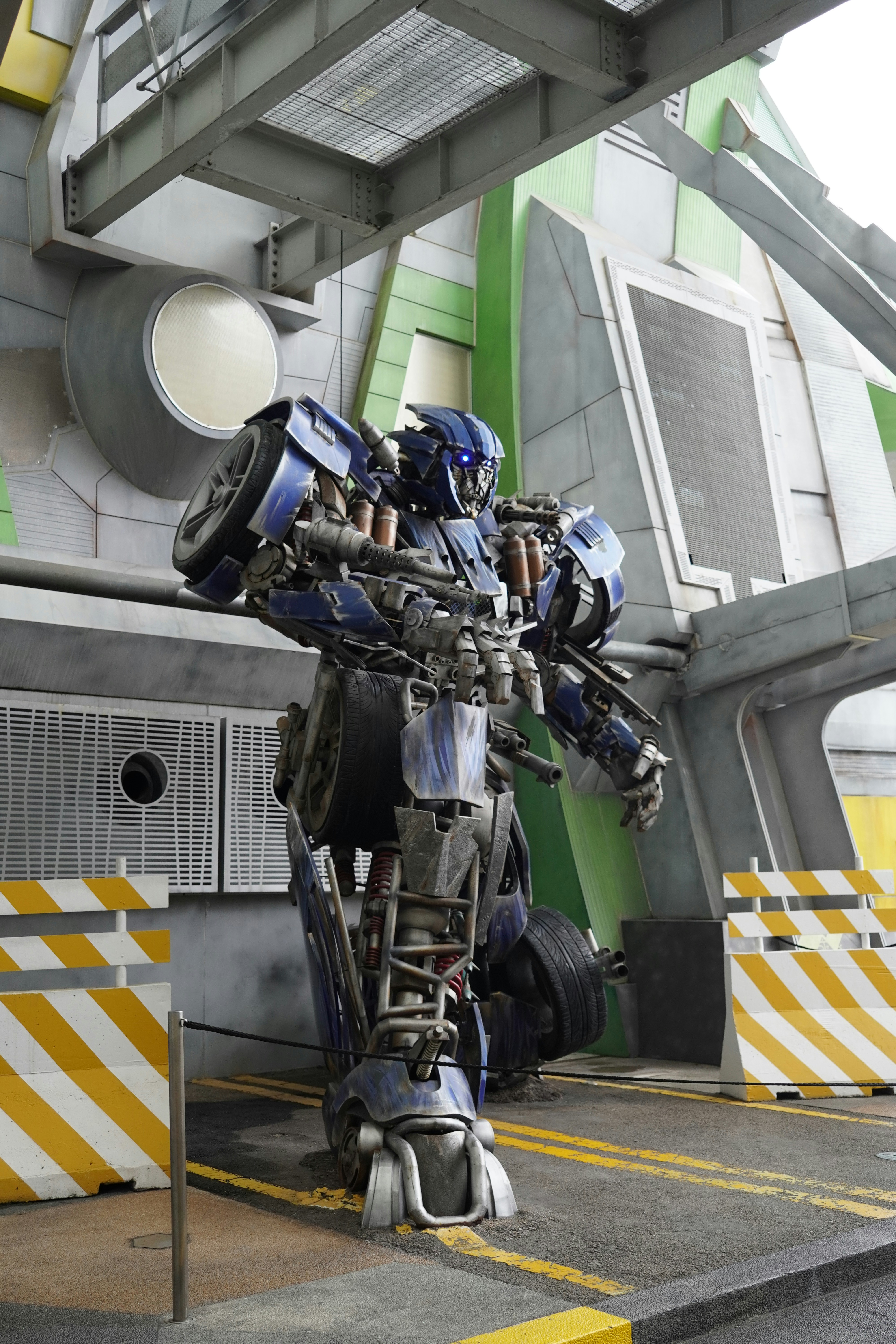 A towering blue robot stands in a futuristic industrial setting with angular green-gray walls and striped barricades in the foreground.