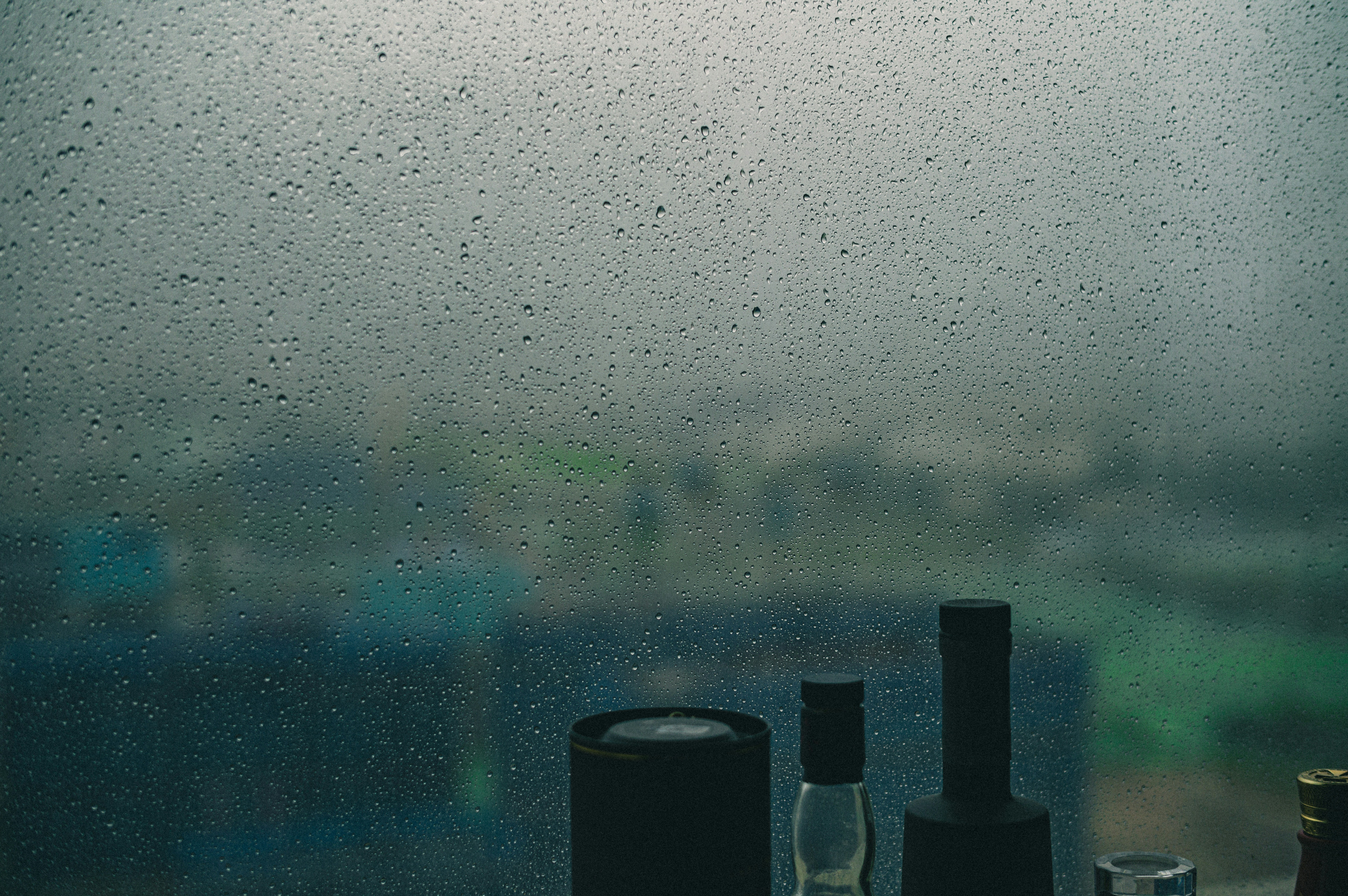 Liquor bottles sit on a windowsill as rain beads the glass, with a softly blurred cityscape beyond.