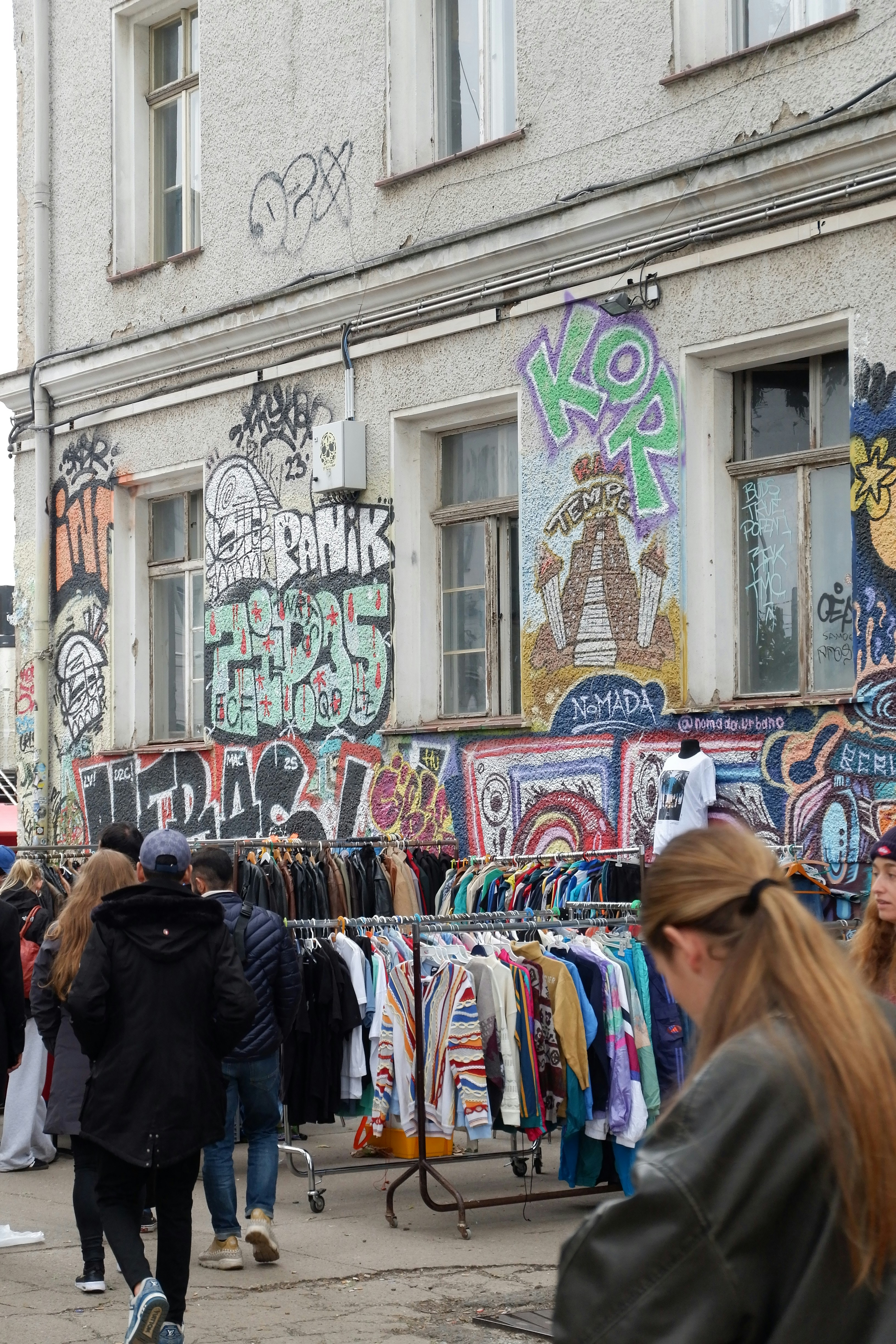 Bustling outdoor market with racks of colorful clothing set against a graffiti-covered wall. Shoppers flank the scene, adding movement to the urban canvas.