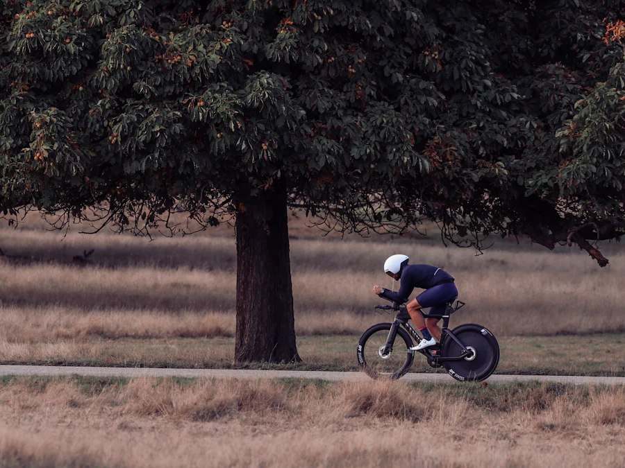 Time-trial cyclist in aerodynamic position riding on a country path past a large oak tree with dry grass in the foreground.