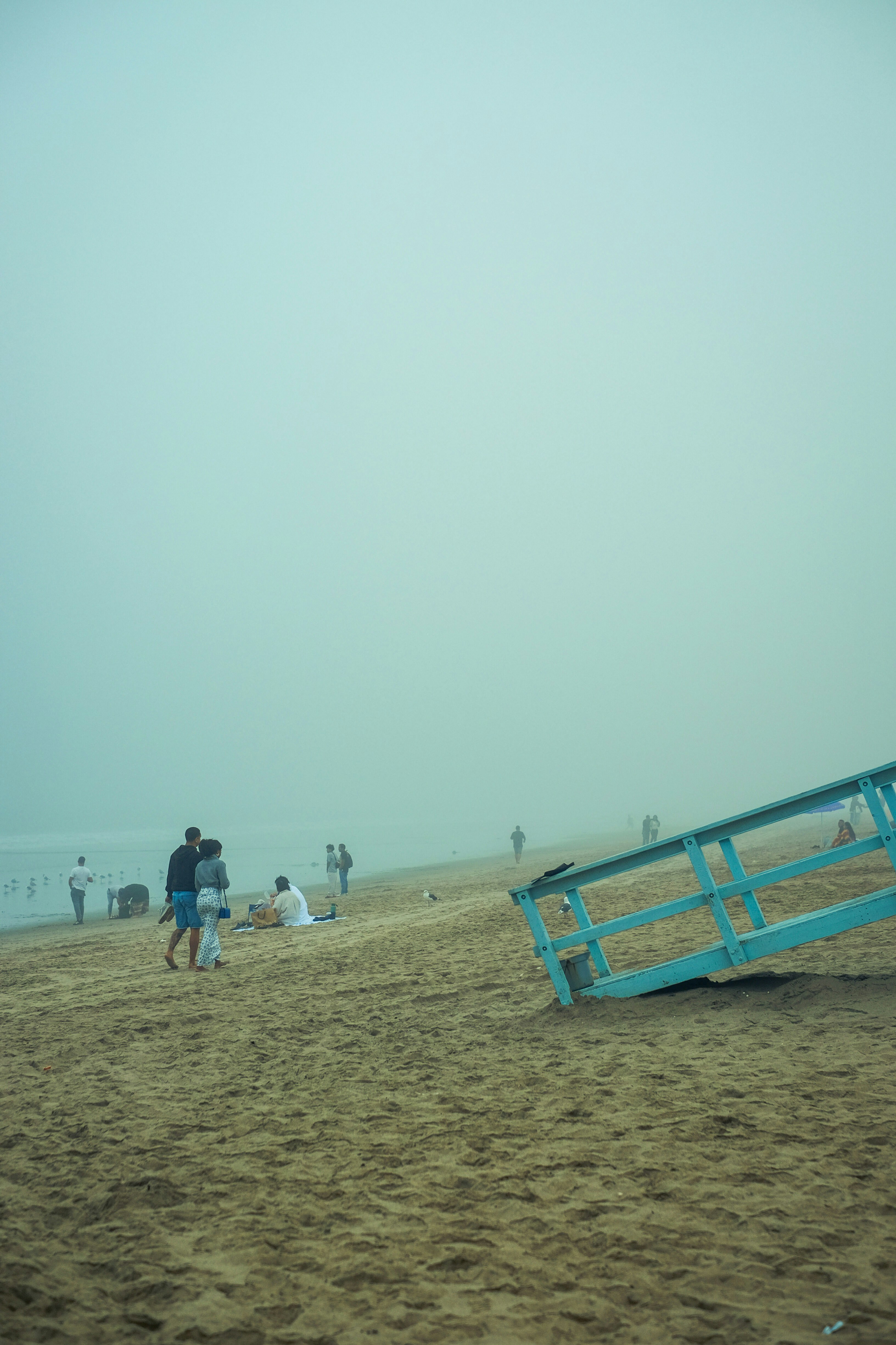Fog-Wrapped Shoreline: A Tilted Blue Lifeguard Chair Observes Quiet Beachgoers