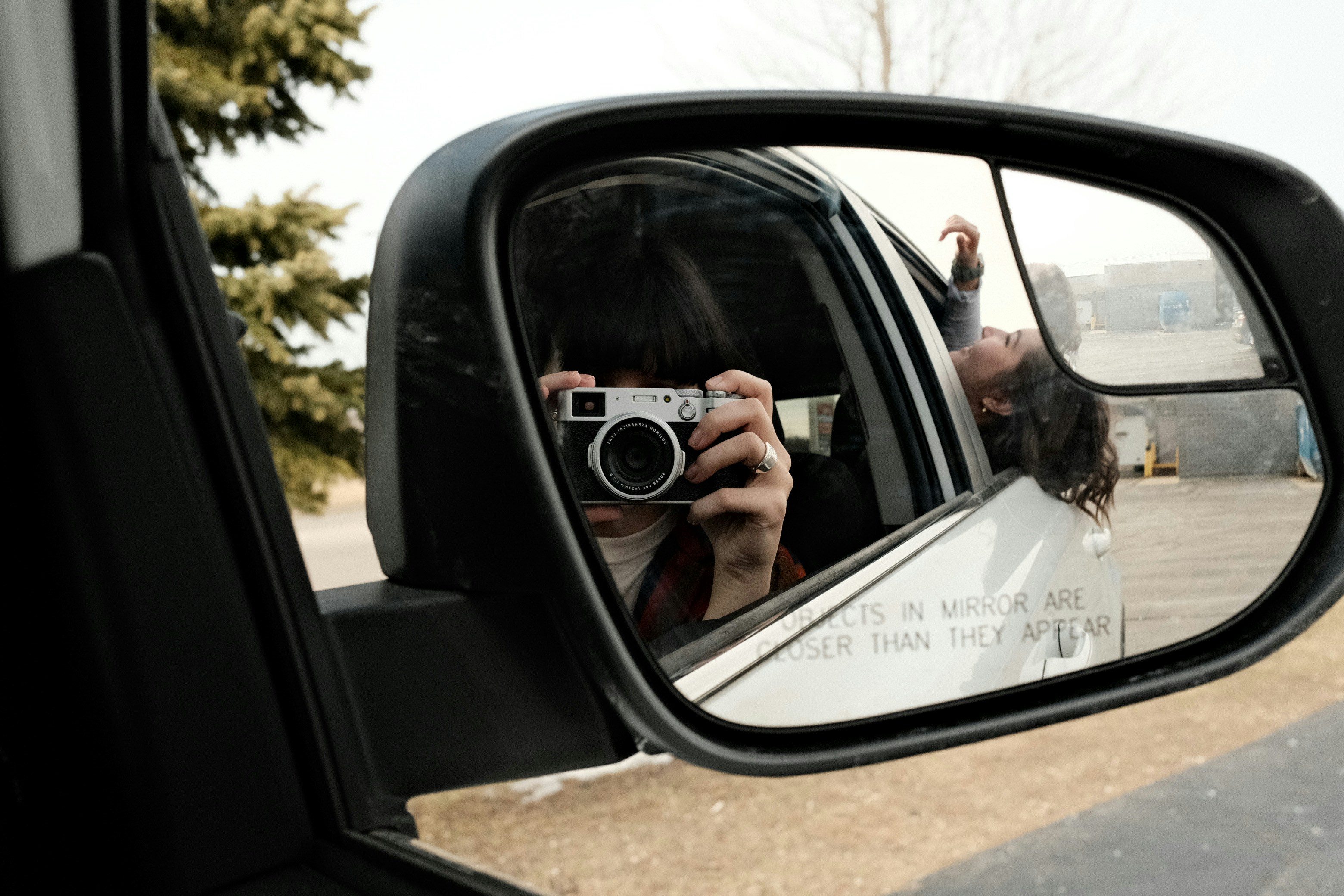 Photographer's reflection in a car side mirror with a playful passenger in the background.