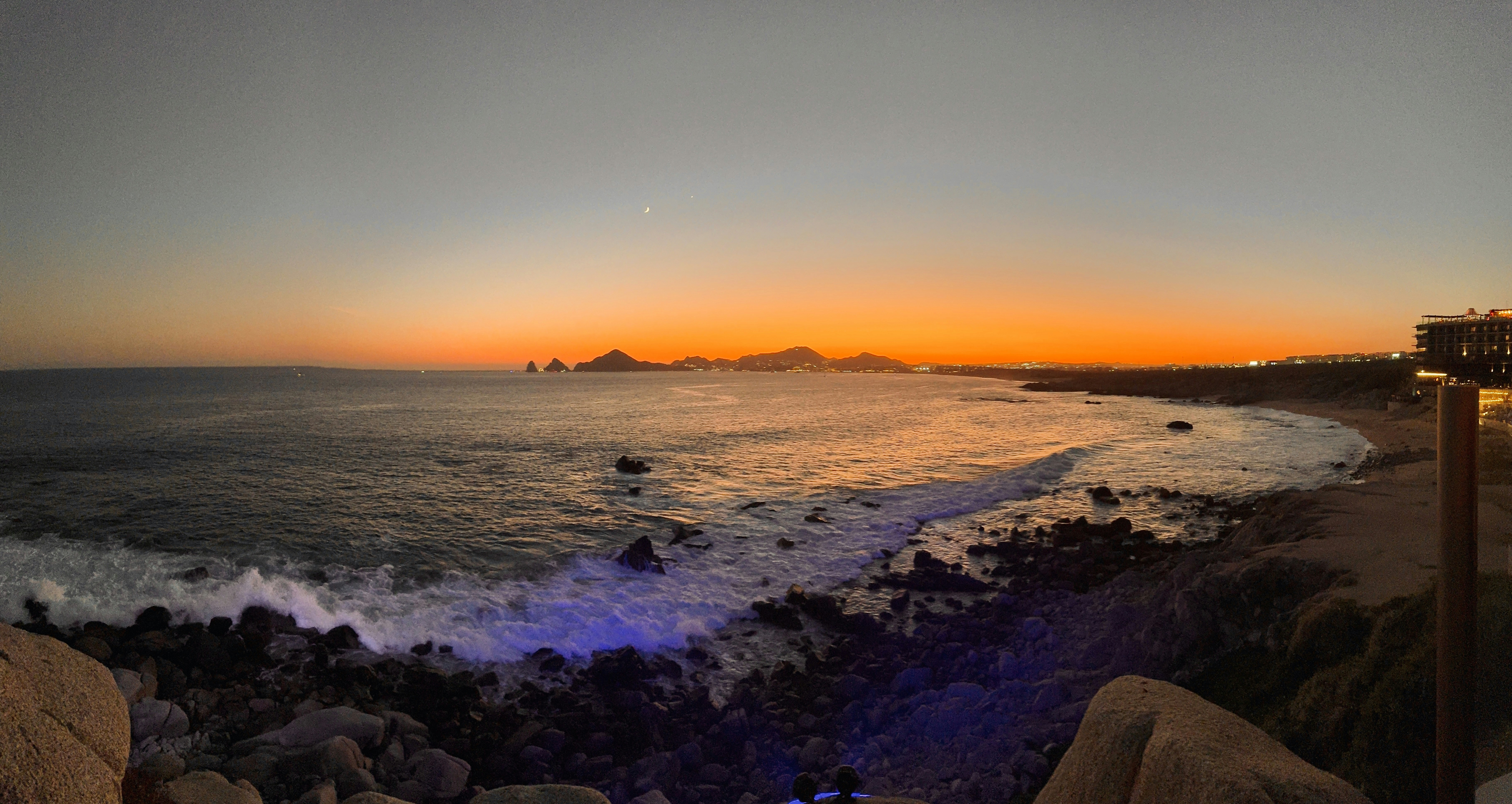 Coastal landscape at dusk with vibrant orange horizon and gentle waves lapping against rocky shore.