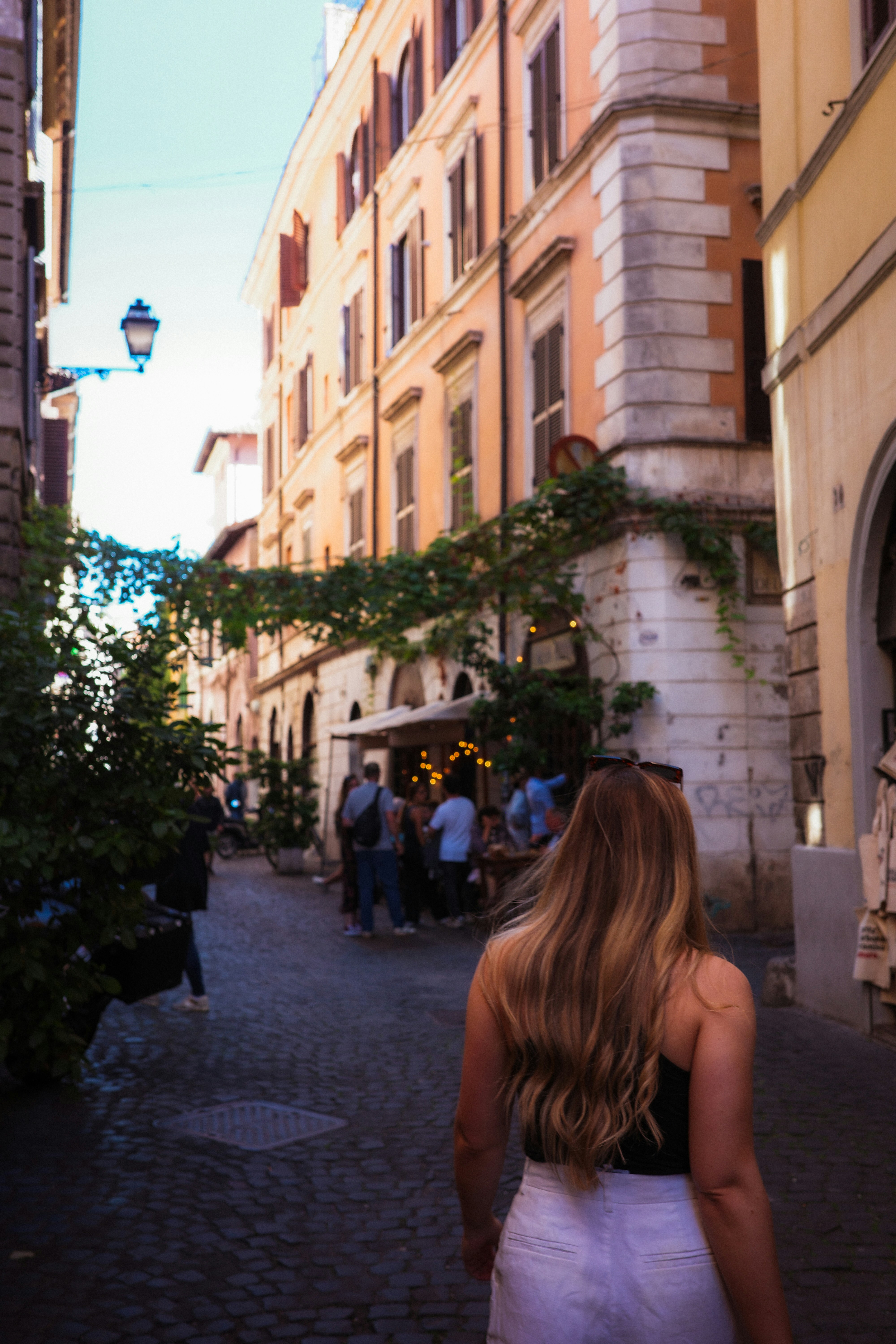 Echoes of a sunlit alley: woman with flowing hair strolling toward a lively courtyard