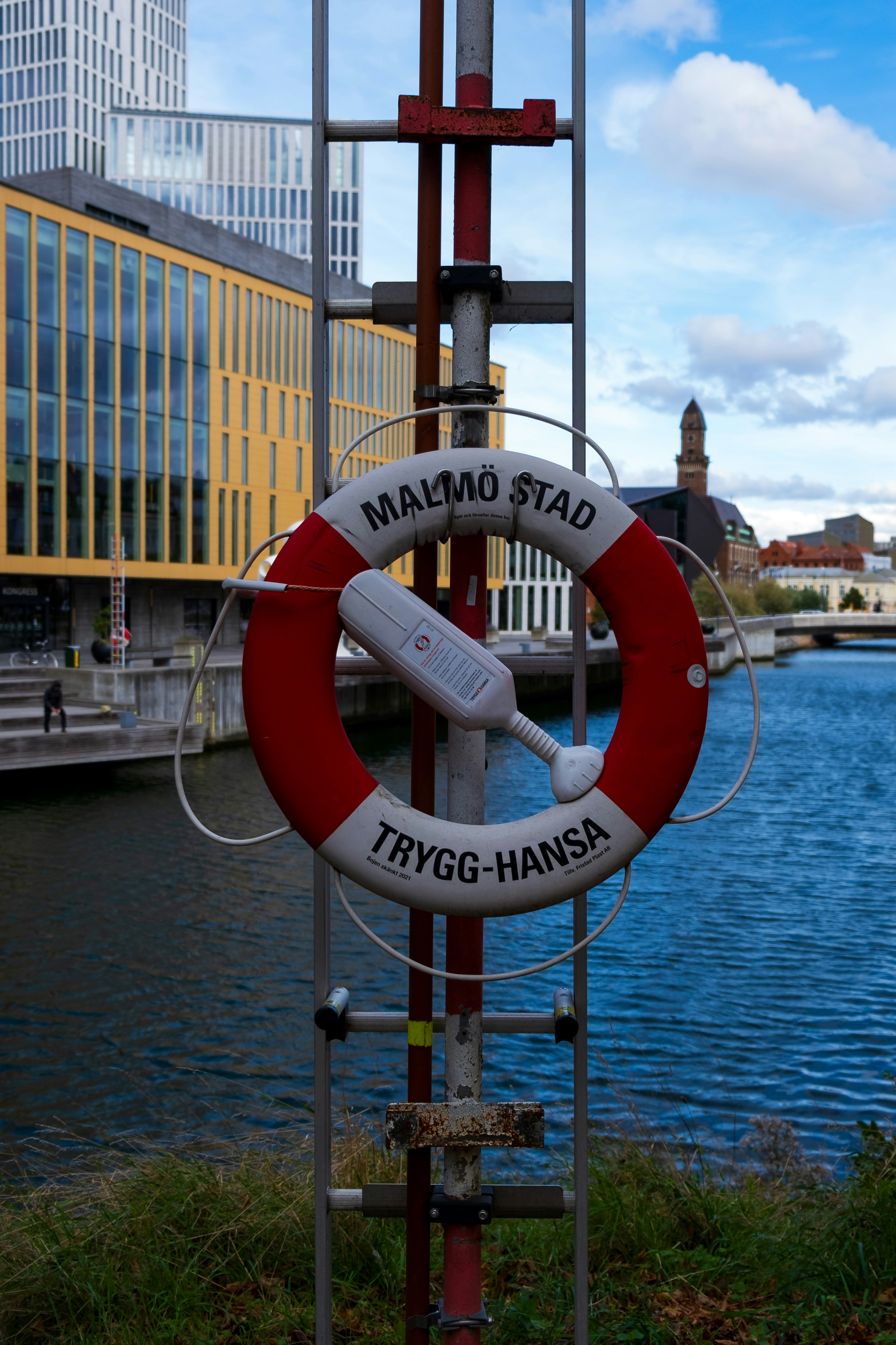 Lifebuoy mounted on a post beside a calm canal, with modern buildings reflecting in the water. A reminder of safety in a vibrant urban setting.
