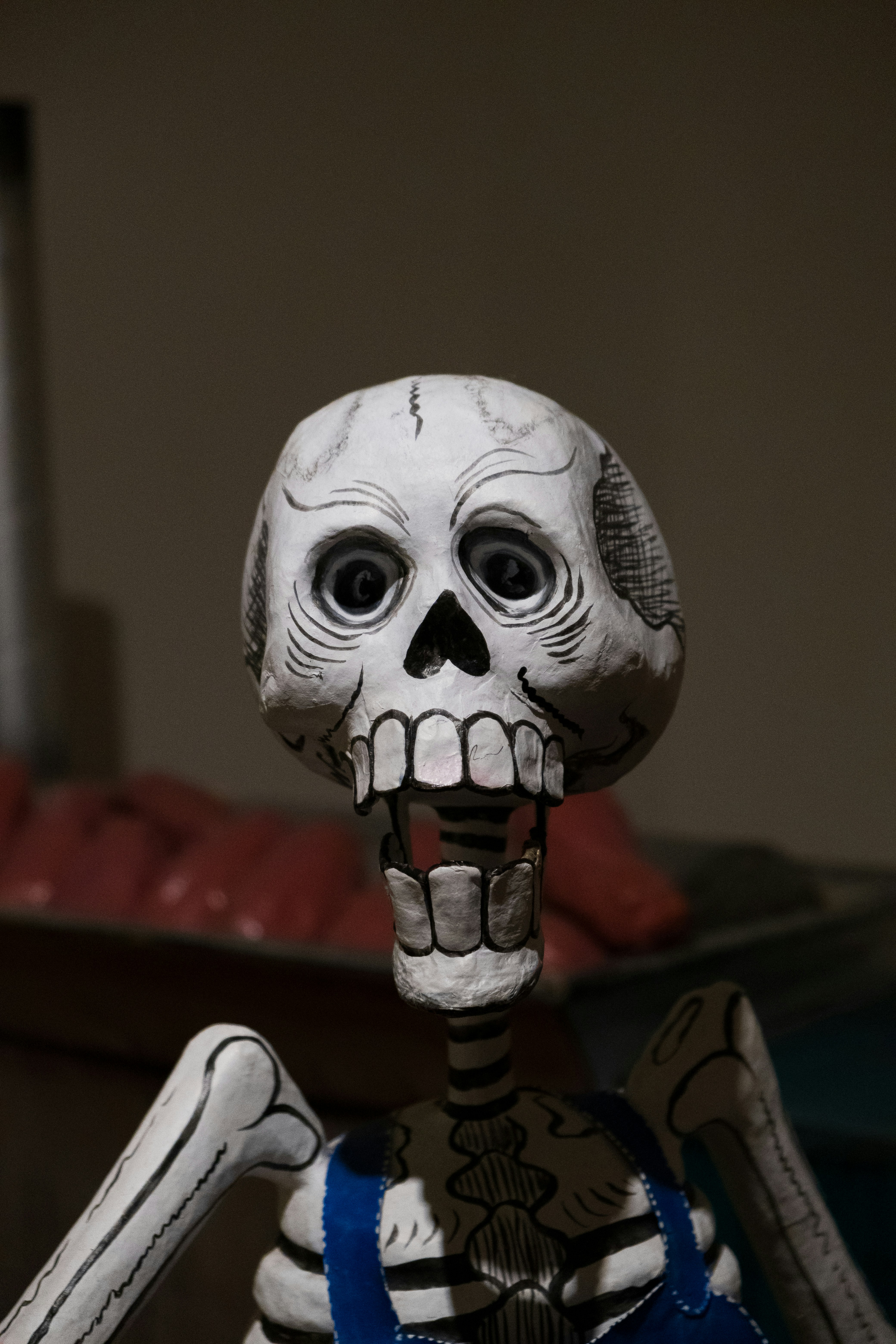 A painted skeleton figure with exaggerated features, wearing a blue harness, stands against a softly blurred background. The playful expression invites curiosity.
