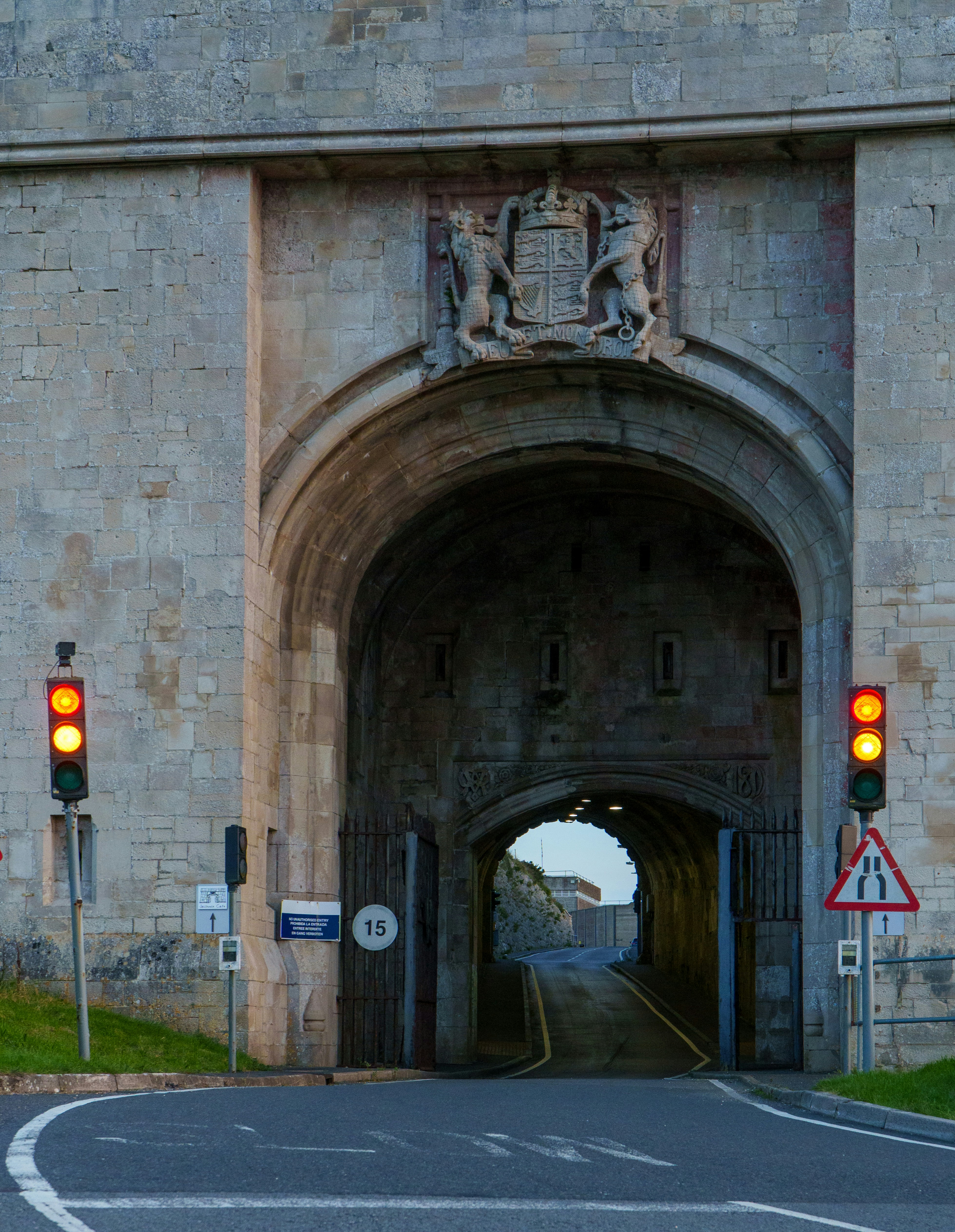 Historic stone archway adorned with a coat of arms, flanked by traffic lights and signage, leading to a winding road beyond.